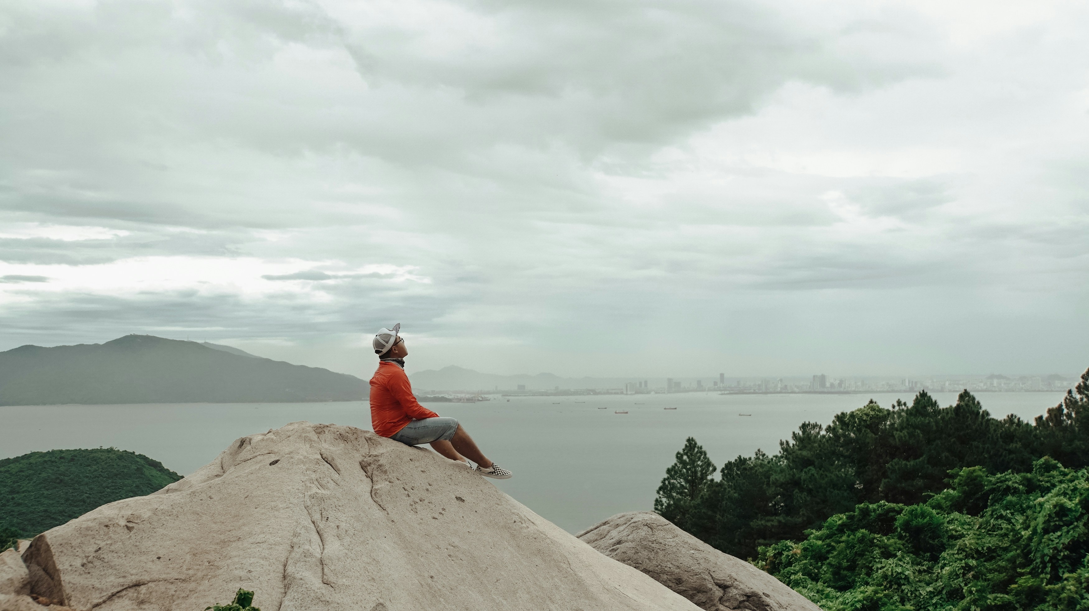A man in an orange shirt sits on a rocky outcrop, gazing over a tranquil seascape under a cloudy sky.