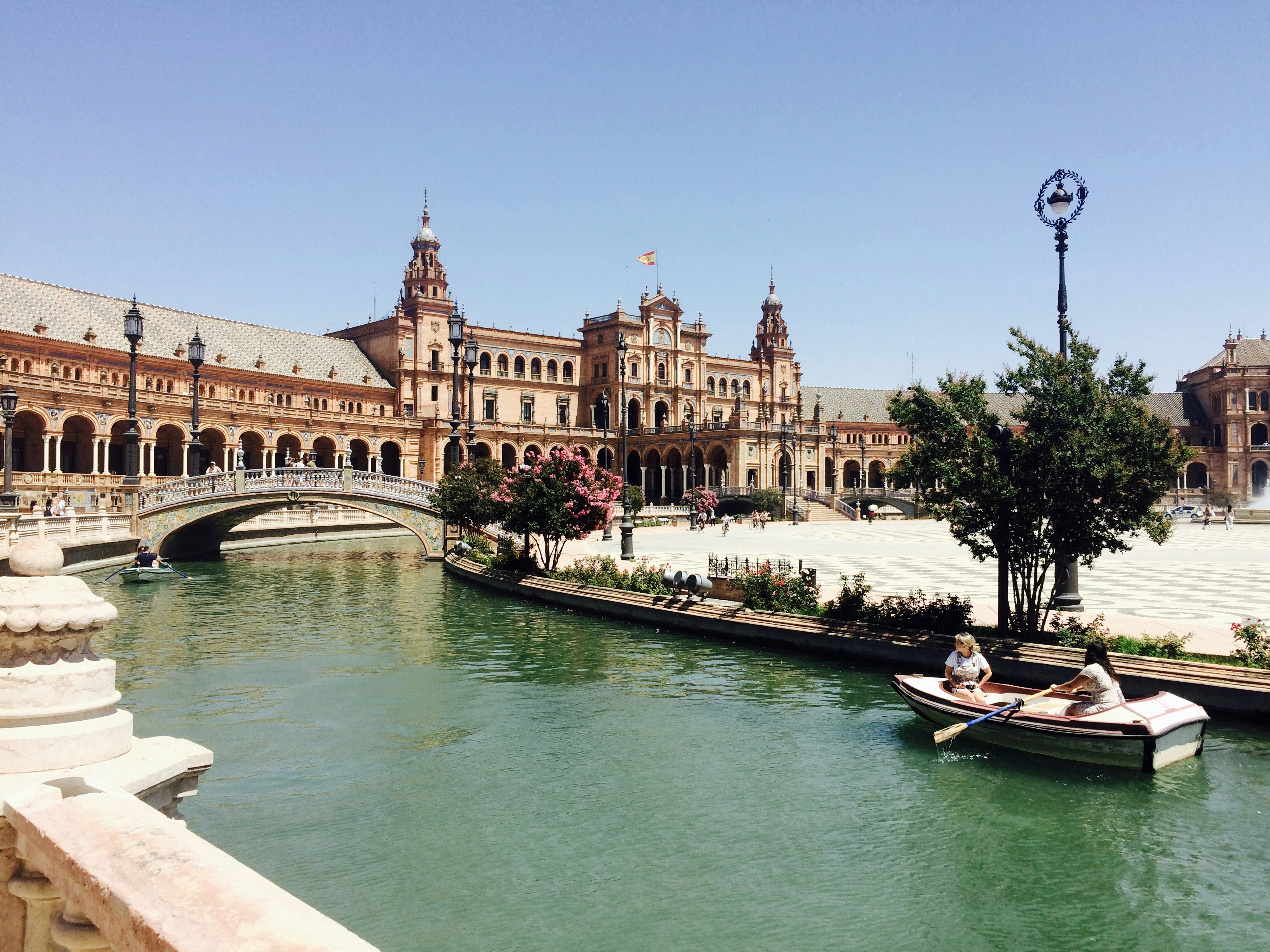 Boaters glide along a tranquil canal by the historic architecture of Plaza de España under a clear blue sky.