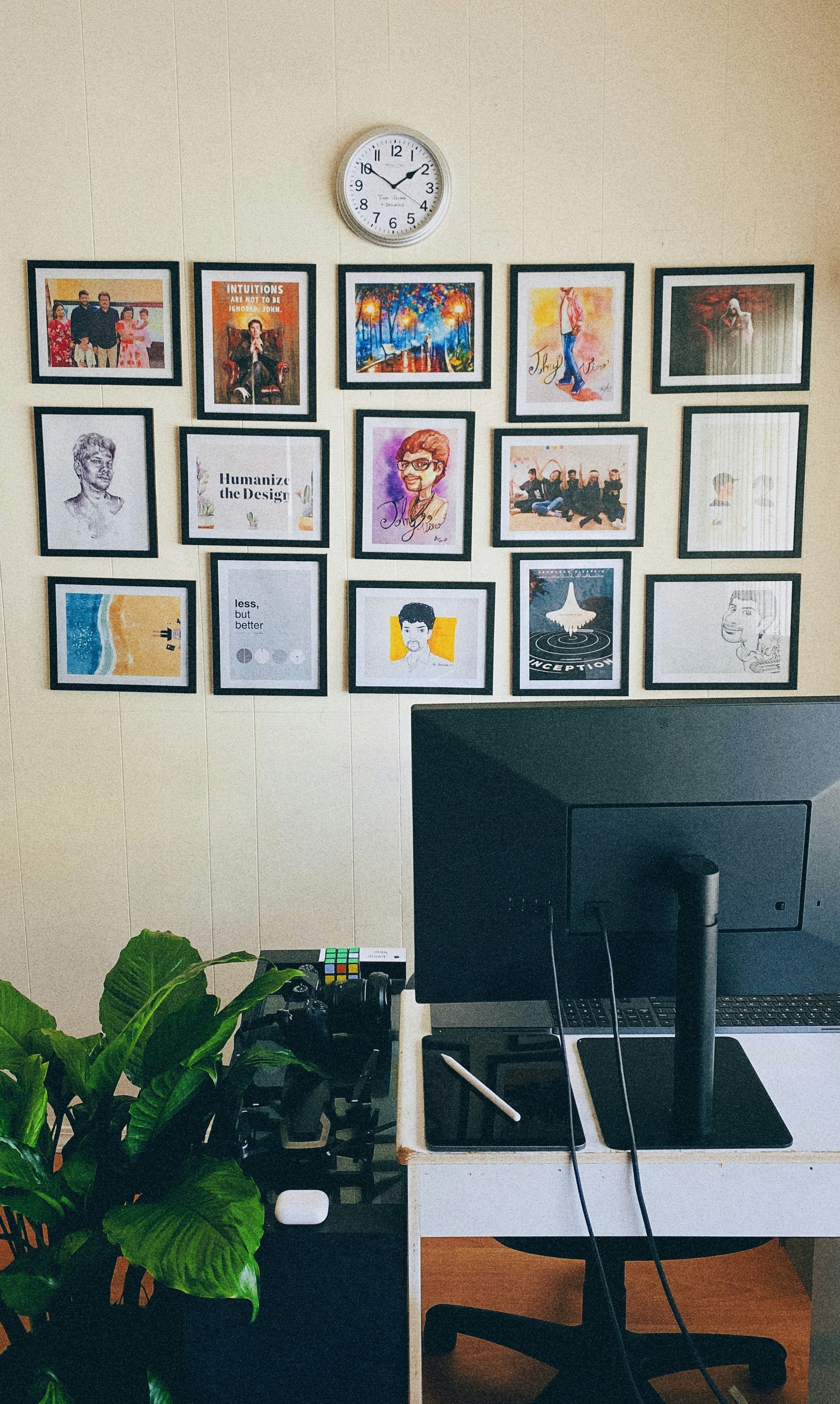 An eclectic display of framed artwork and photographs on a wall, complemented by a desk setup featuring a computer and a vibrant plant.