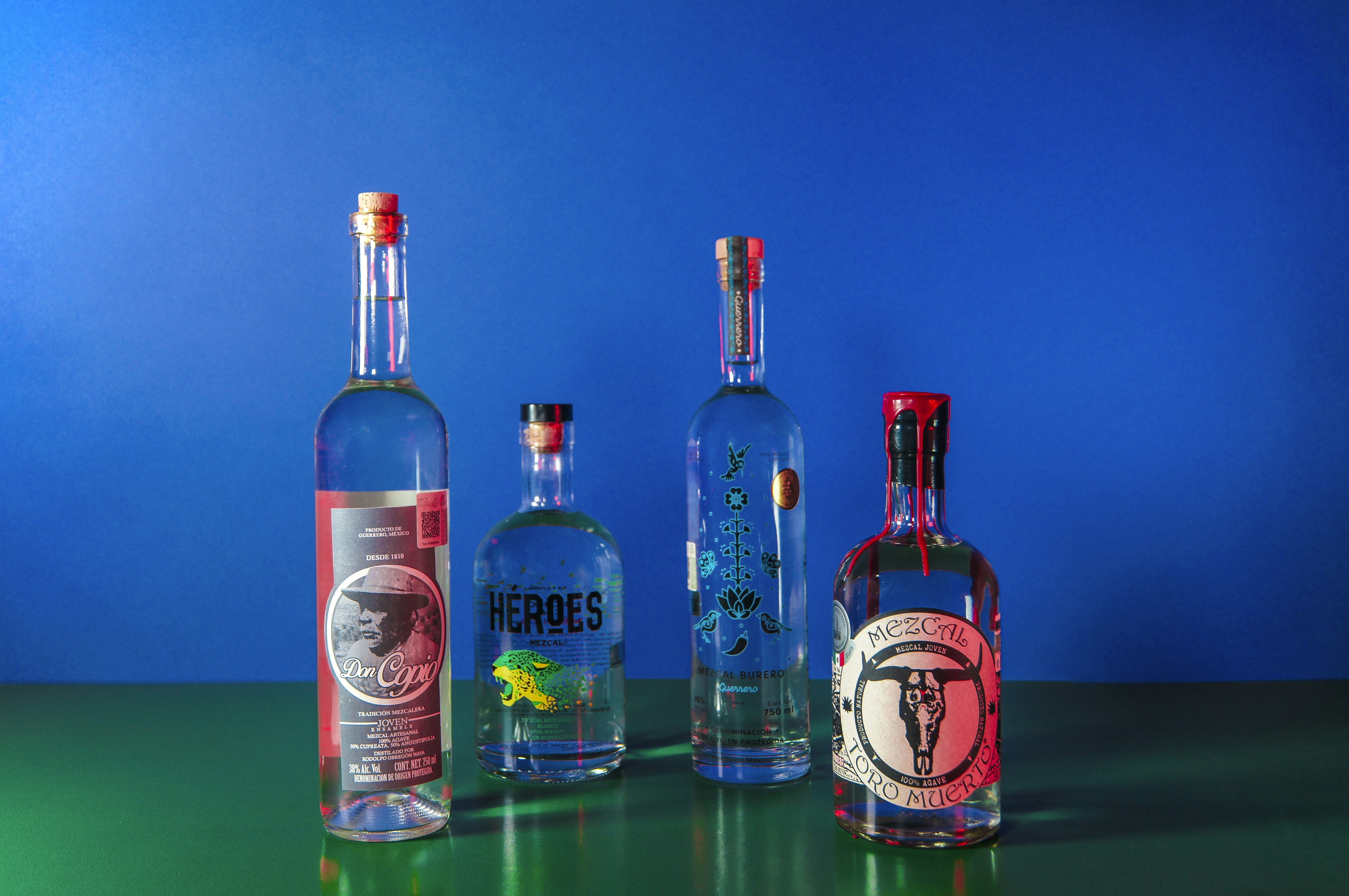 Four mezcal bottles arranged on a reflective green surface against a vivid blue backdrop.