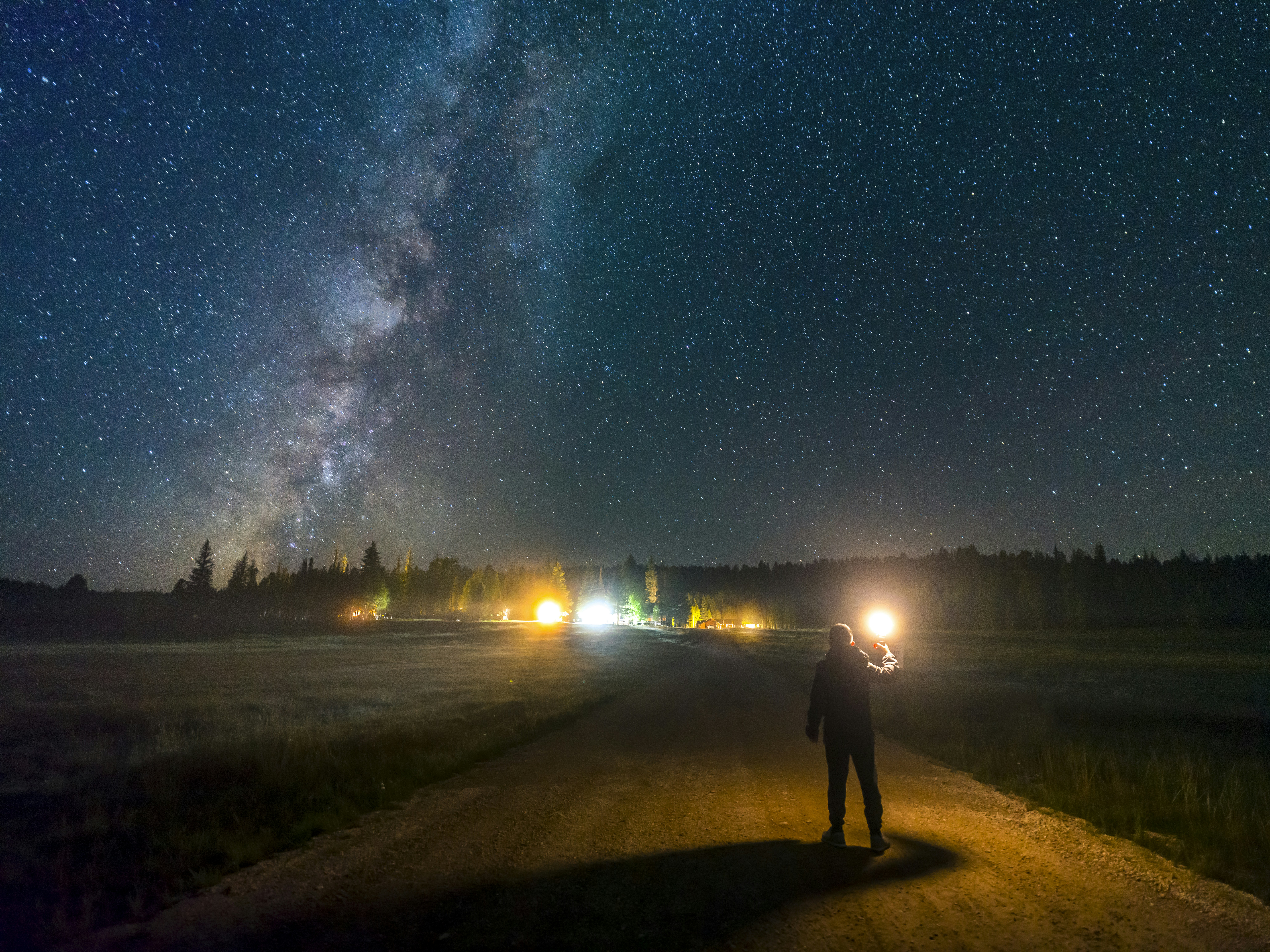 A lone figure stands on a gravel path illuminated by a flashlight, gazing up at the sprawling Milky Way amidst a star-filled sky.