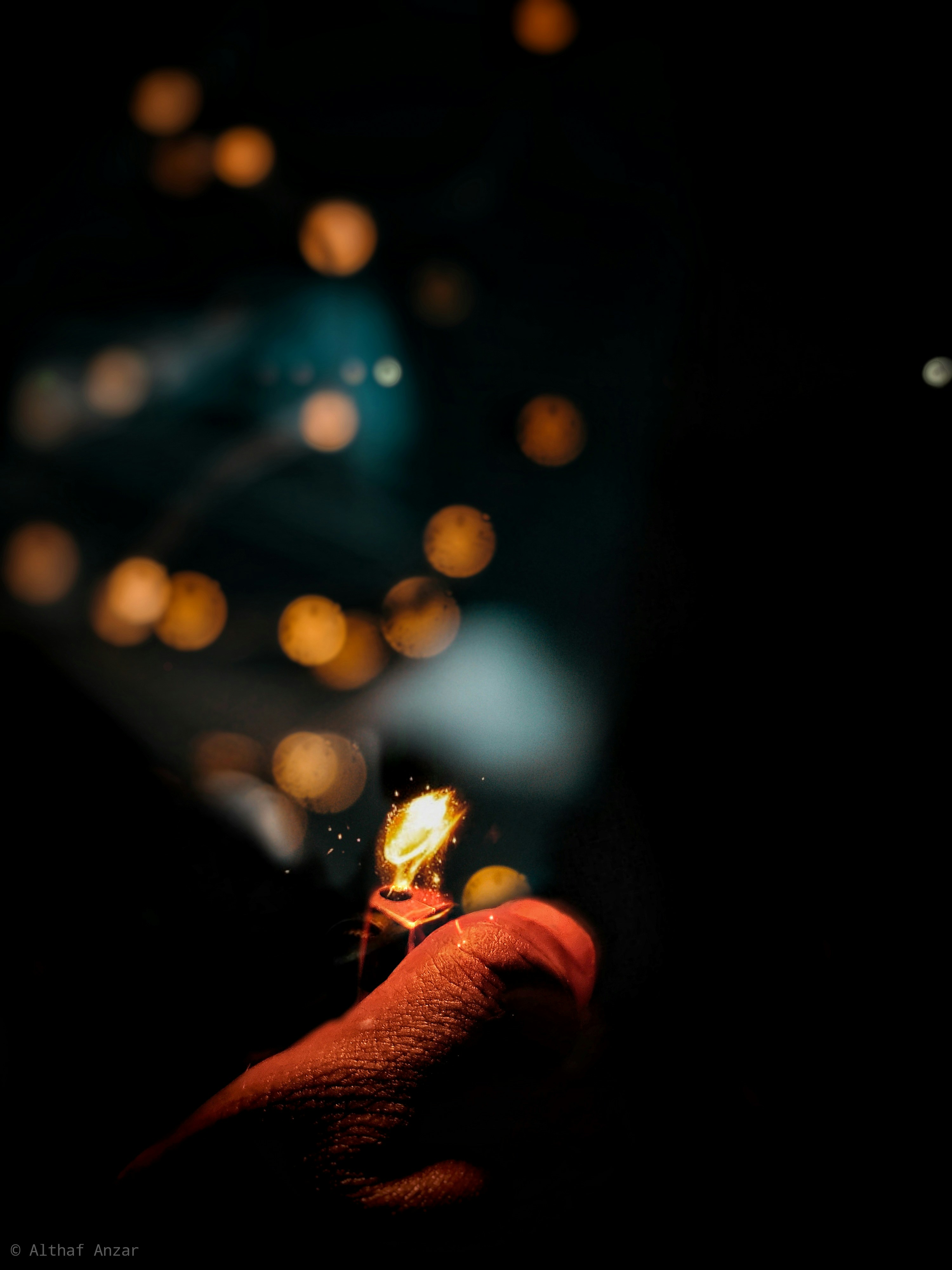Close-up photograph of a small flame lit by a lighter held by a finger. Warm bokeh orbs add a dreamlike glow in the dark background.