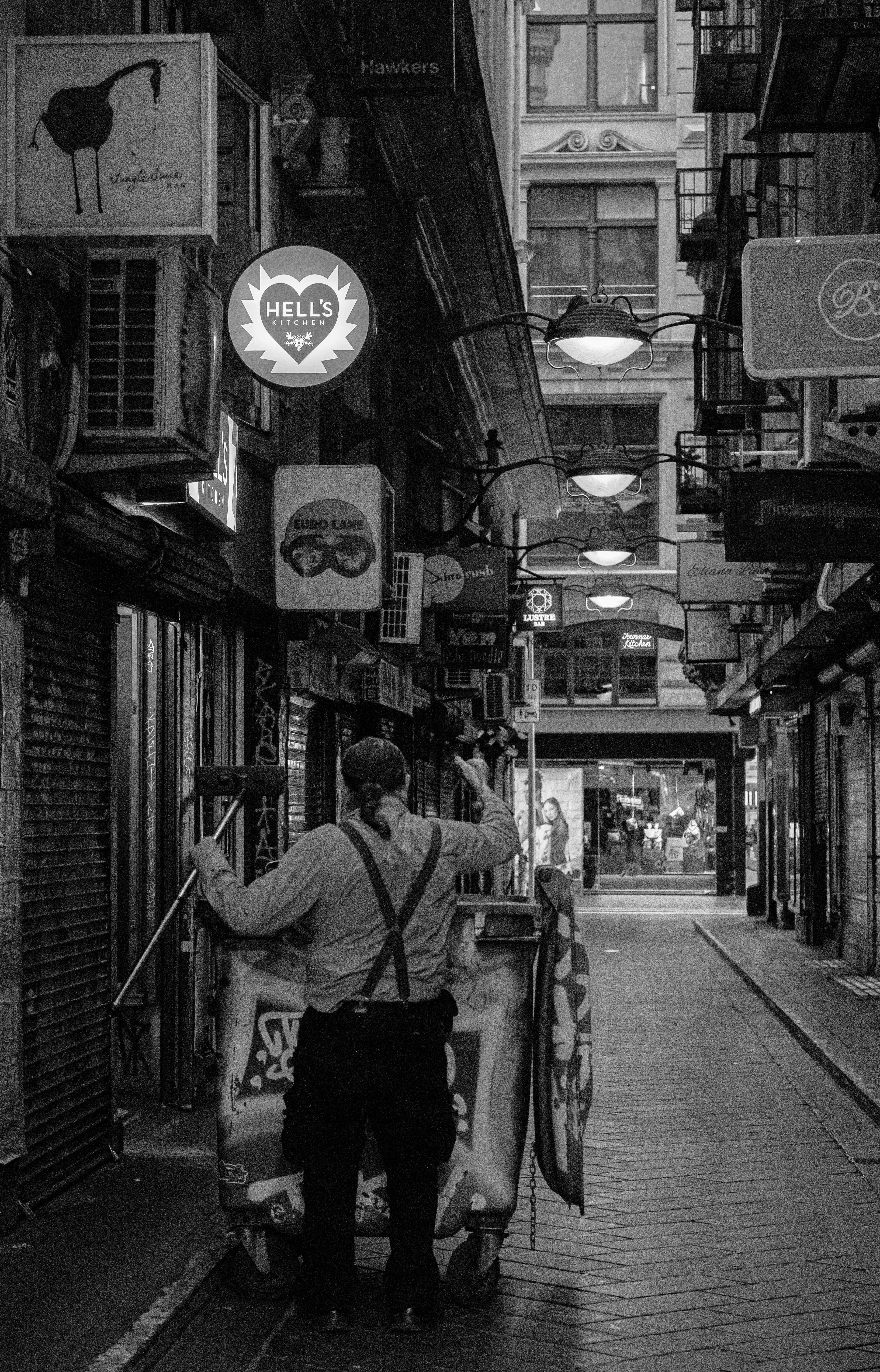 A street worker pushes a cart through a narrow alley adorned with vibrant signage, capturing the essence of urban life. The scene is rendered in black and white, highlighting the contrast between light and shadow.
