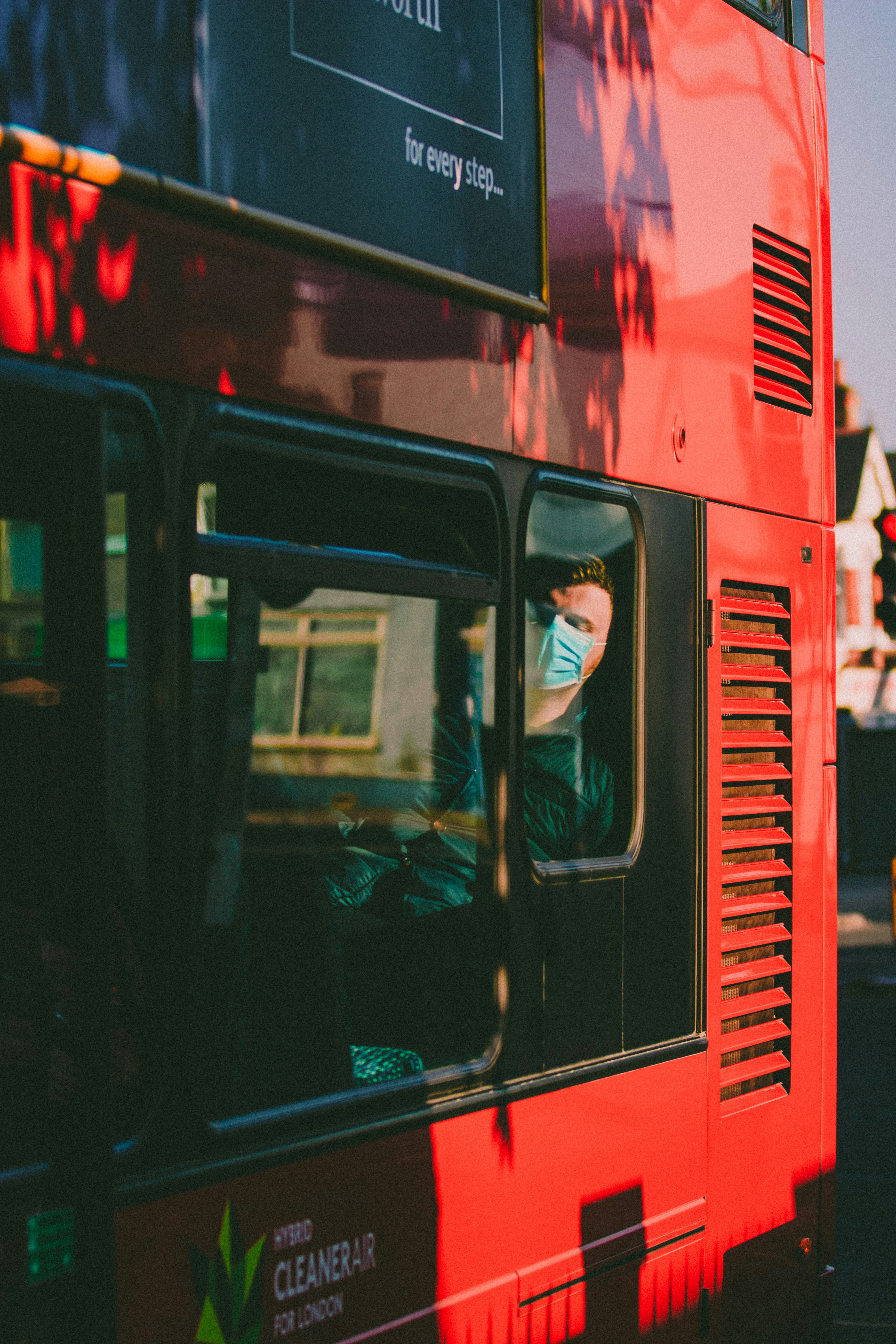 A person wearing a mask gazes out from a red London bus, framed by the vibrant colors and shadows of the urban environment.