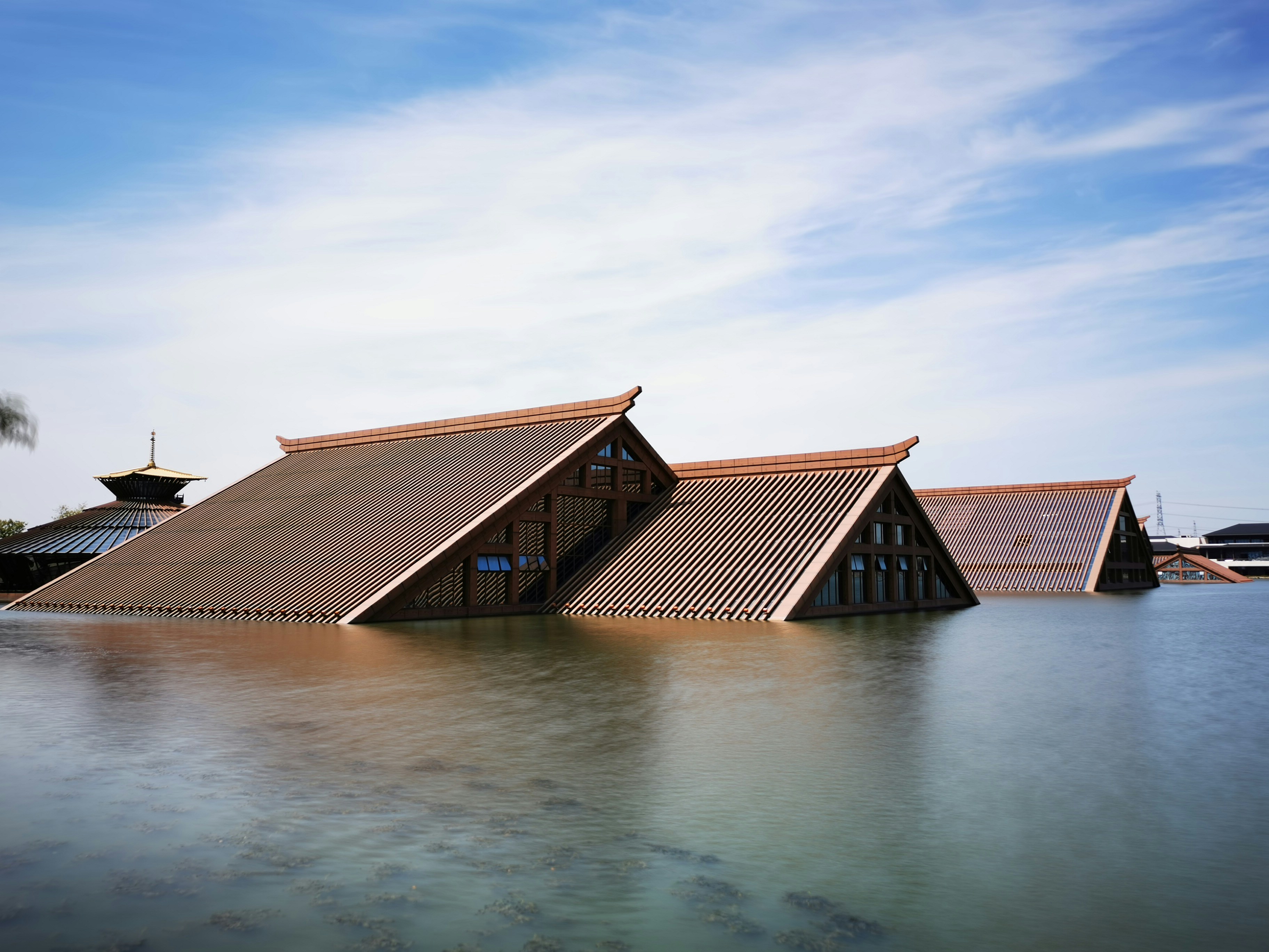 Triangular-roof structures rise from a calm lagoon, their bases submerged and reflections softened by the still water under a clear sky.