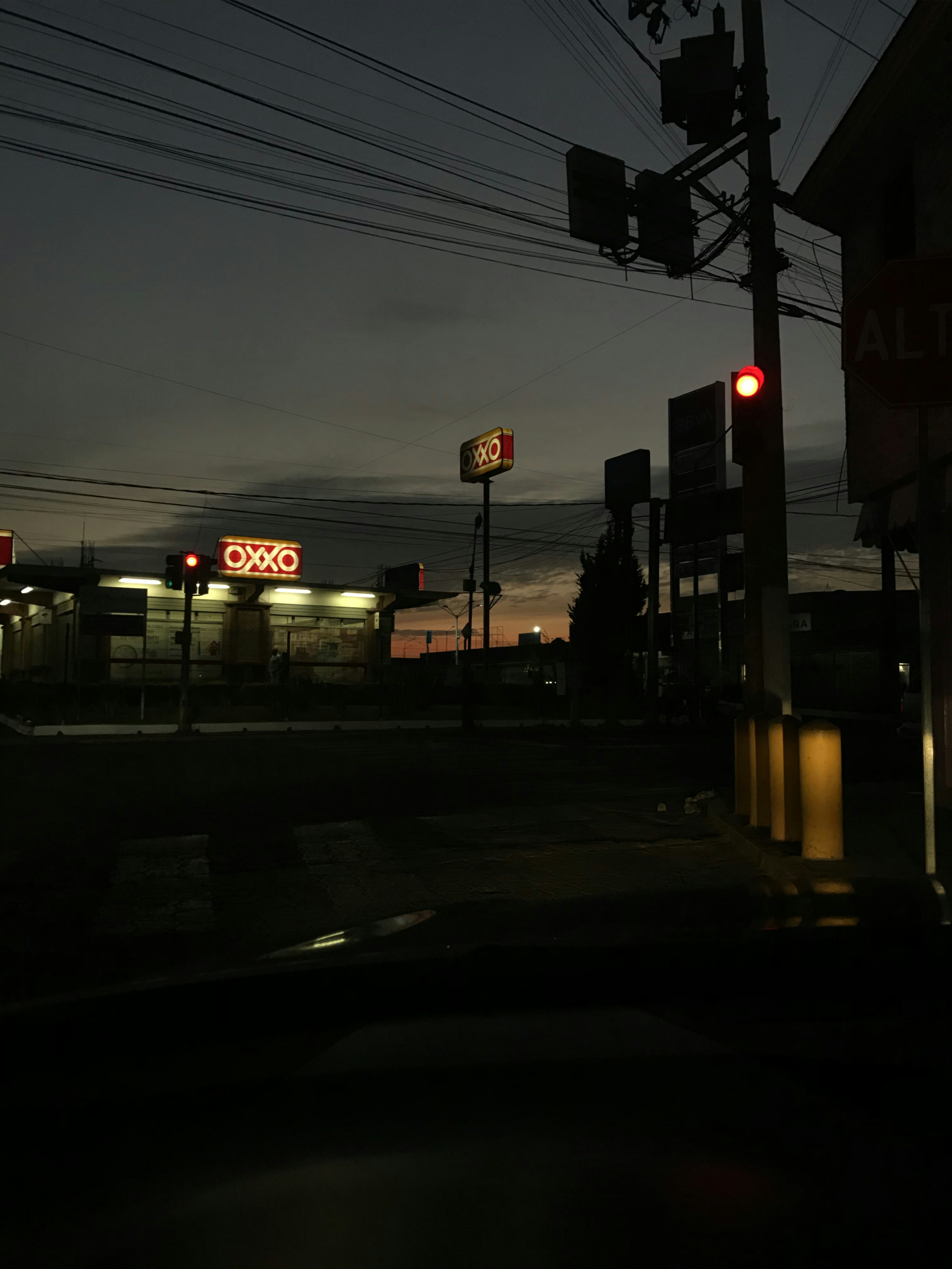 OXXO convenience store illuminated against a twilight sky, with traffic lights and power lines framing the scene. The atmosphere captures the transition from day to night.