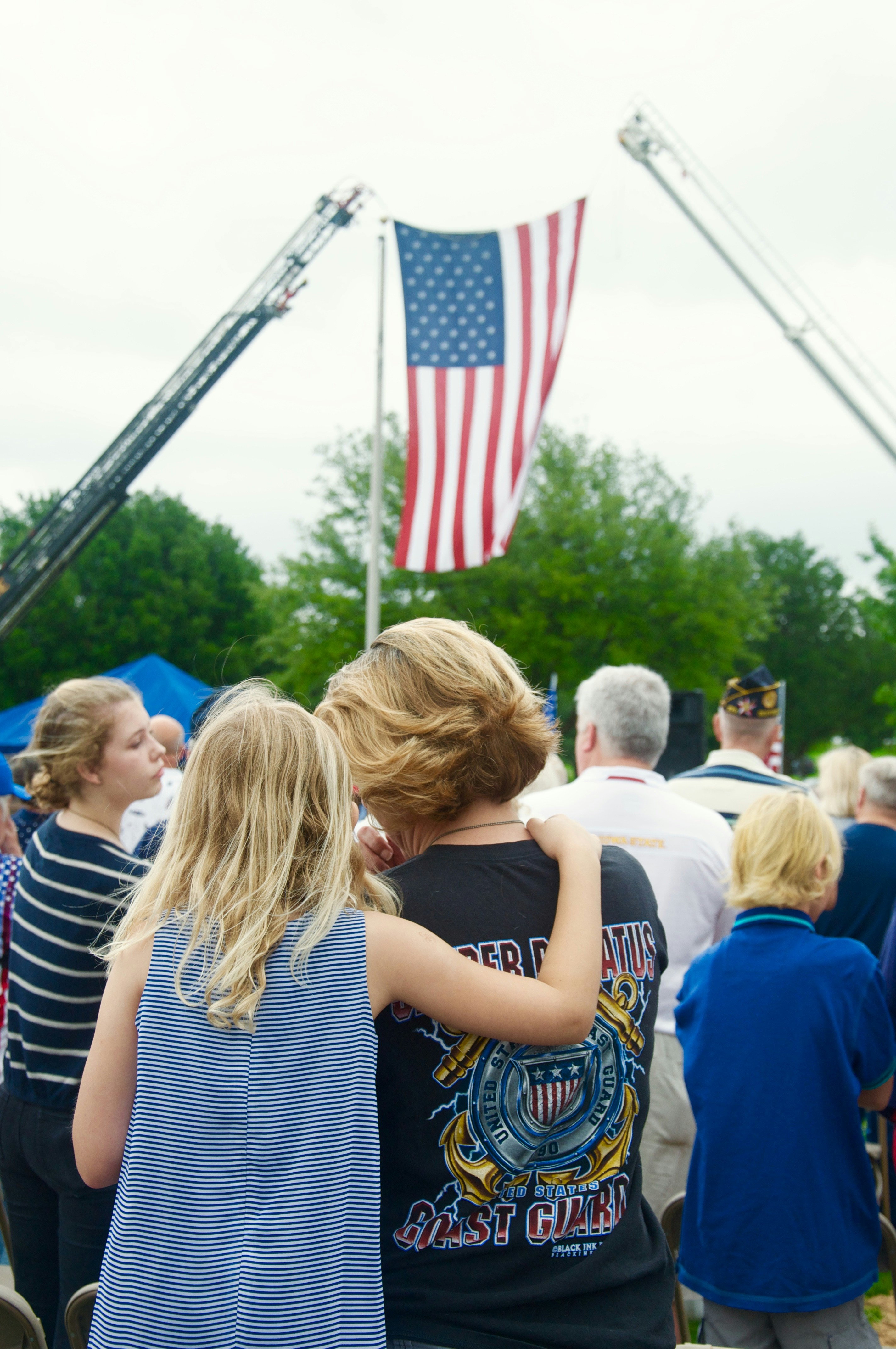 Two children share a heartfelt embrace while watching a patriotic event, with a large American flag displayed prominently in the background.