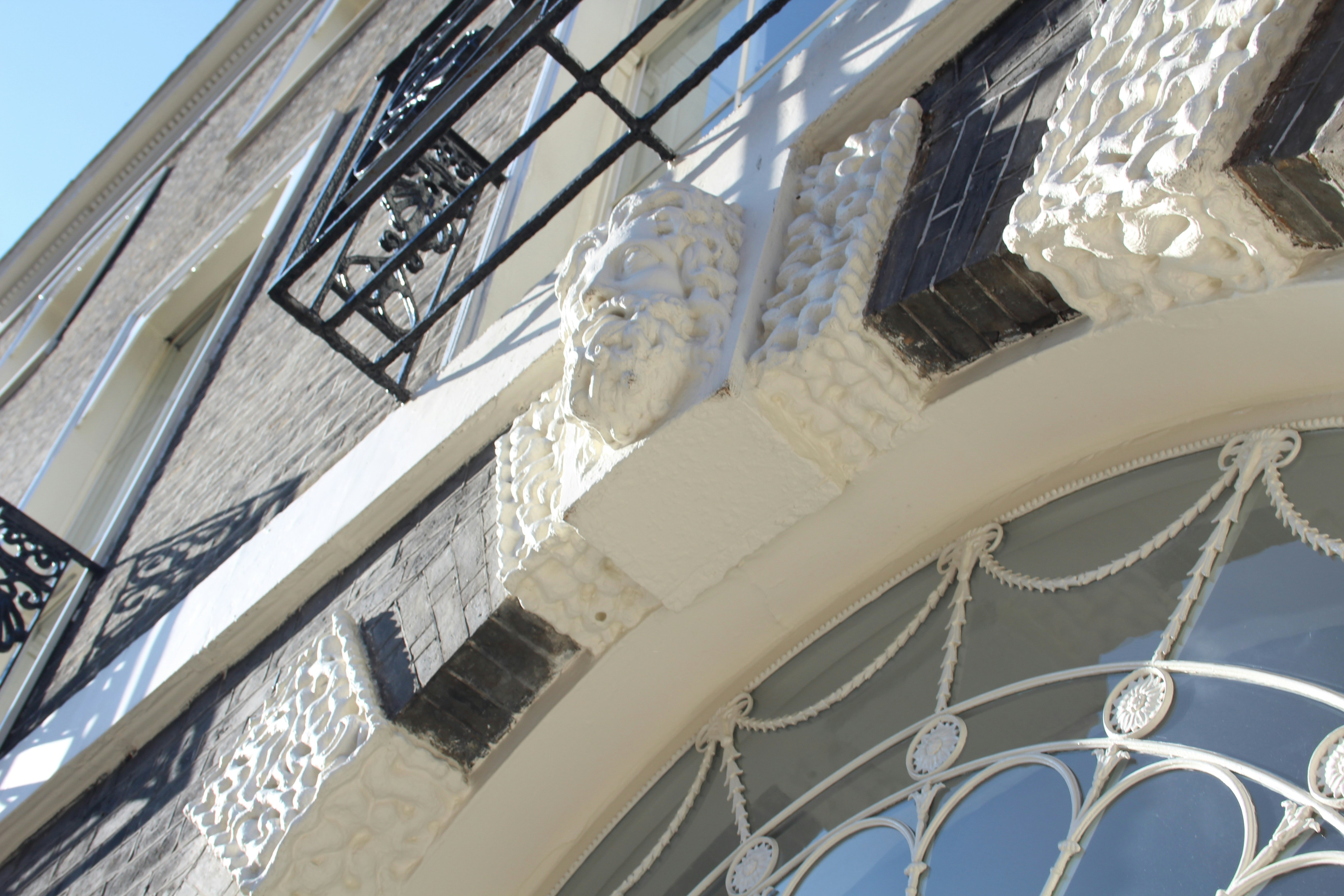 Intricate architectural details featuring a sculpted face atop an ornate archway, framed by elegant wrought iron balconies. The sunlight accentuates the textures and craftsmanship.