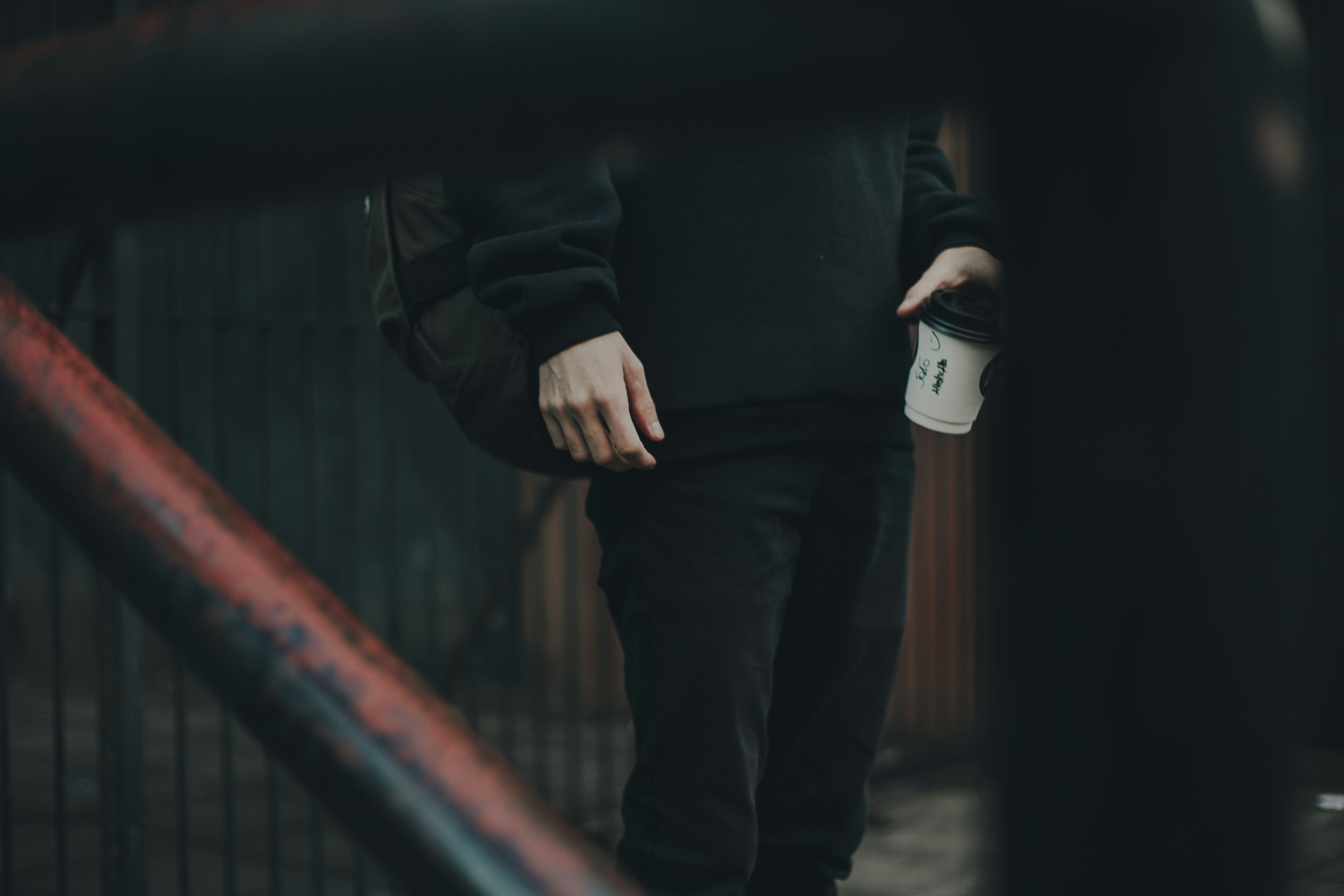 Person in black clothing holding a white disposable cup, partially obscured by an urban railing.