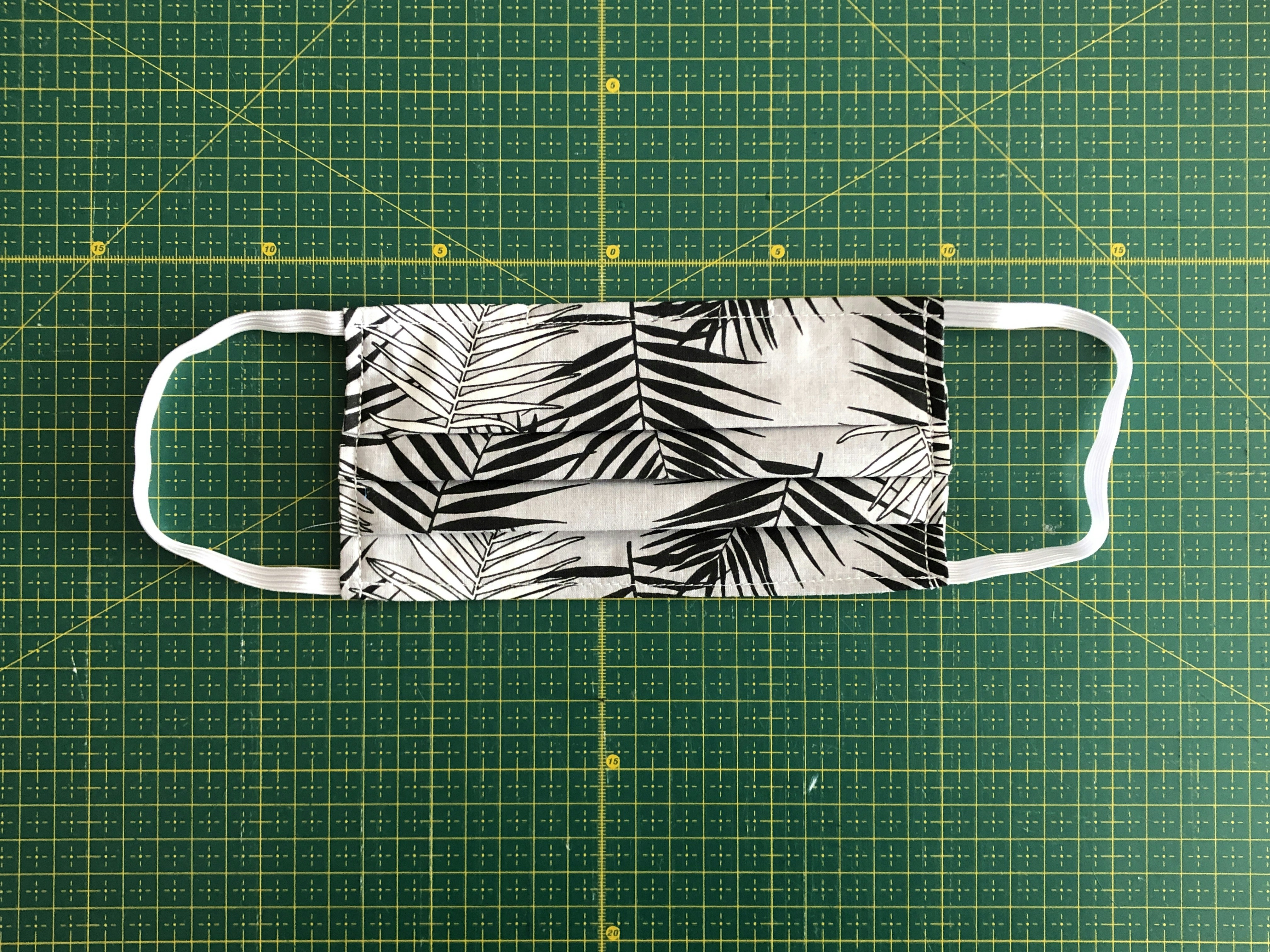 Stylish fabric mask featuring a black and white palm leaf design, centered on a green cutting mat with grid lines.