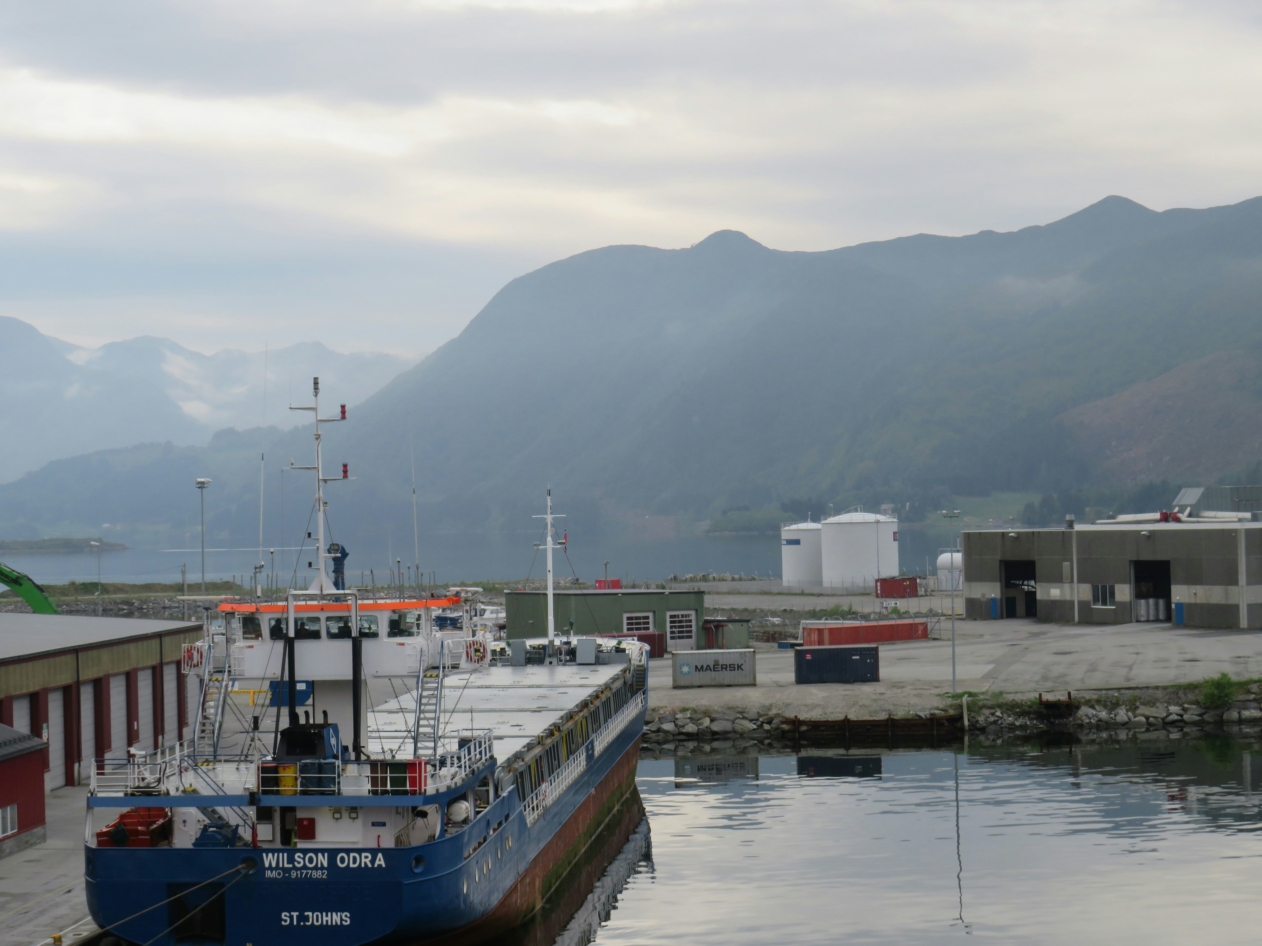 Cargo ship Wilson Odra docked in a tranquil harbor, surrounded by mountains and industrial buildings. The calm waters reflect the serene landscape.
