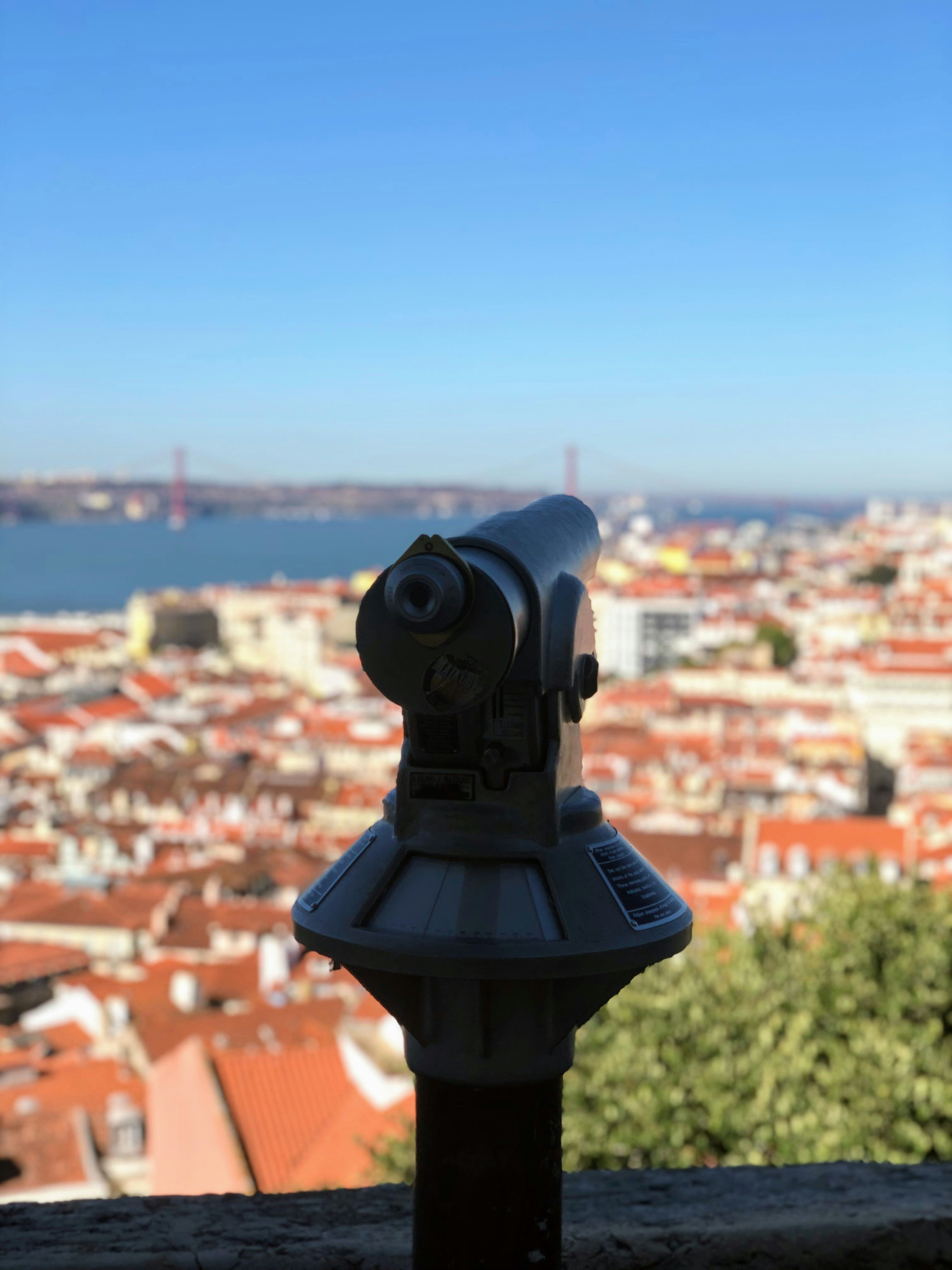 Viewing telescope overlooking a vibrant cityscape with red rooftops and a distant bridge. The clear blue sky enhances the panoramic view.