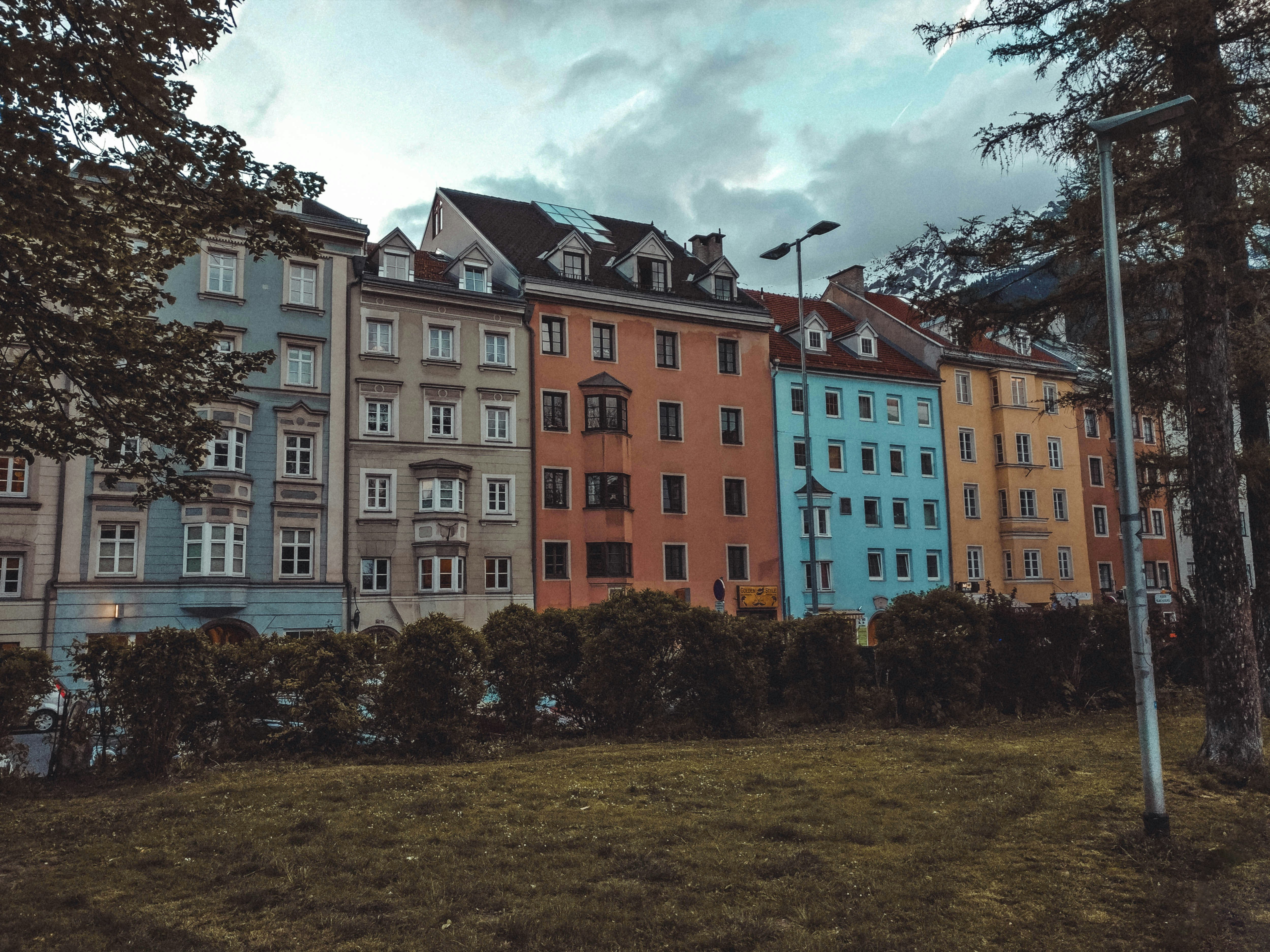 Colorful townhouses stand in a row beneath an overcast sky, framed by trees and a grassy foreground.