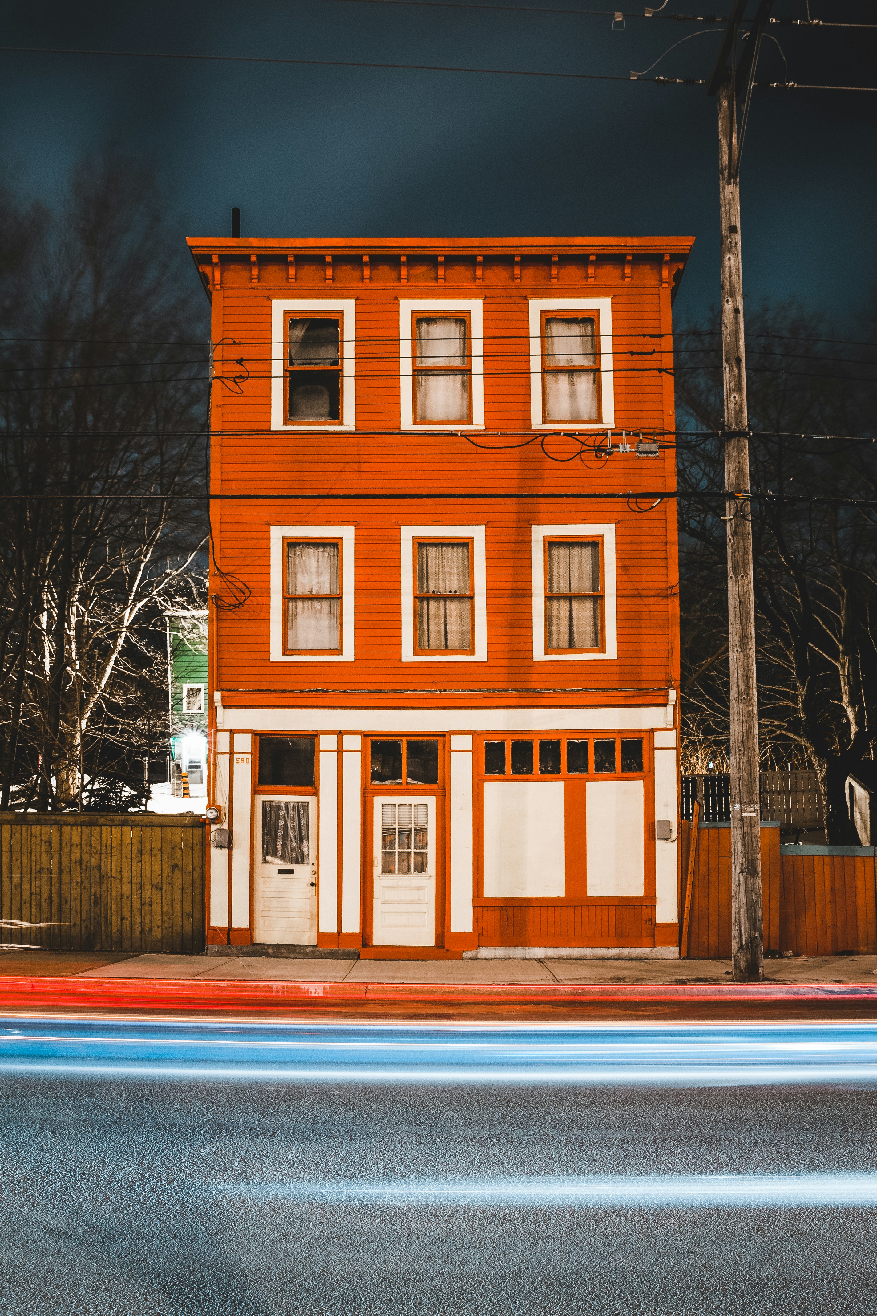 Bright orange building stands against a night sky, with light trails from passing vehicles creating a dynamic contrast. The scene captures the essence of urban energy.