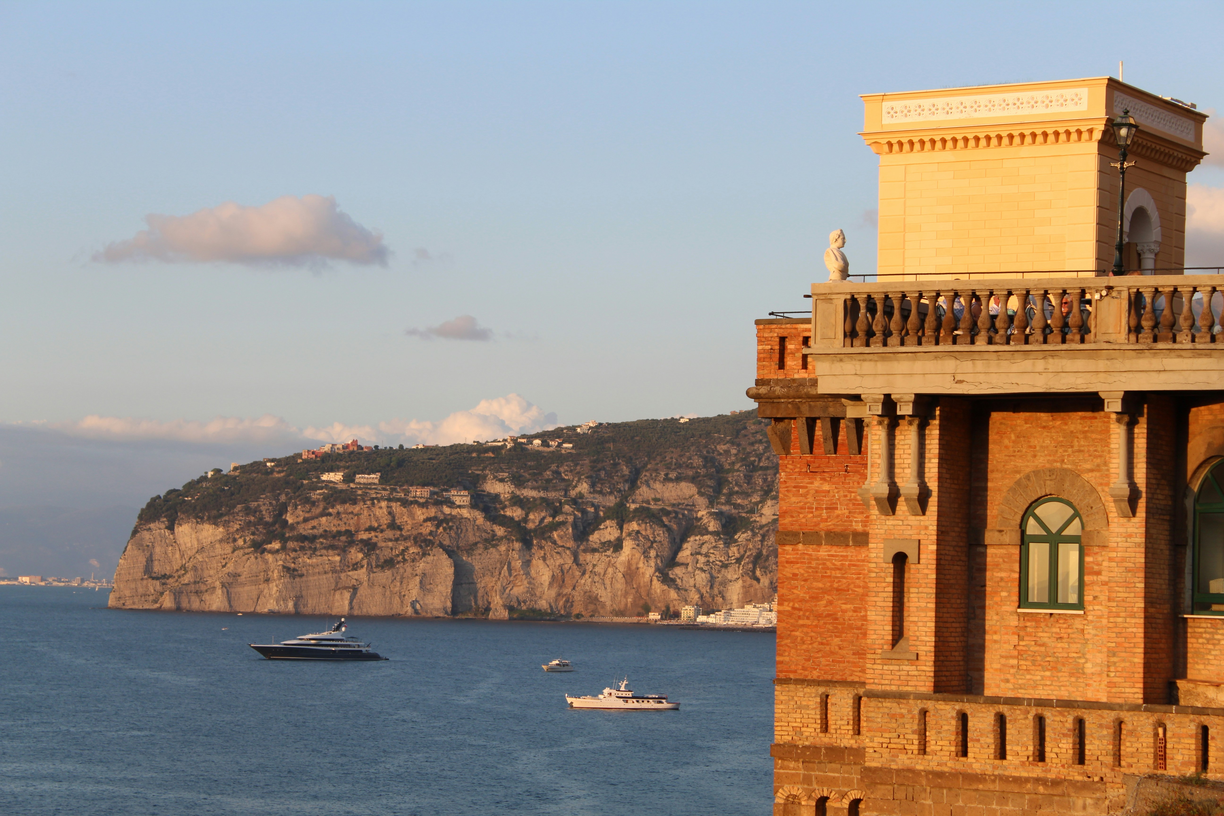 Brown stone building overlooks a calm sea with distant cliffs and boats under a clear sky.