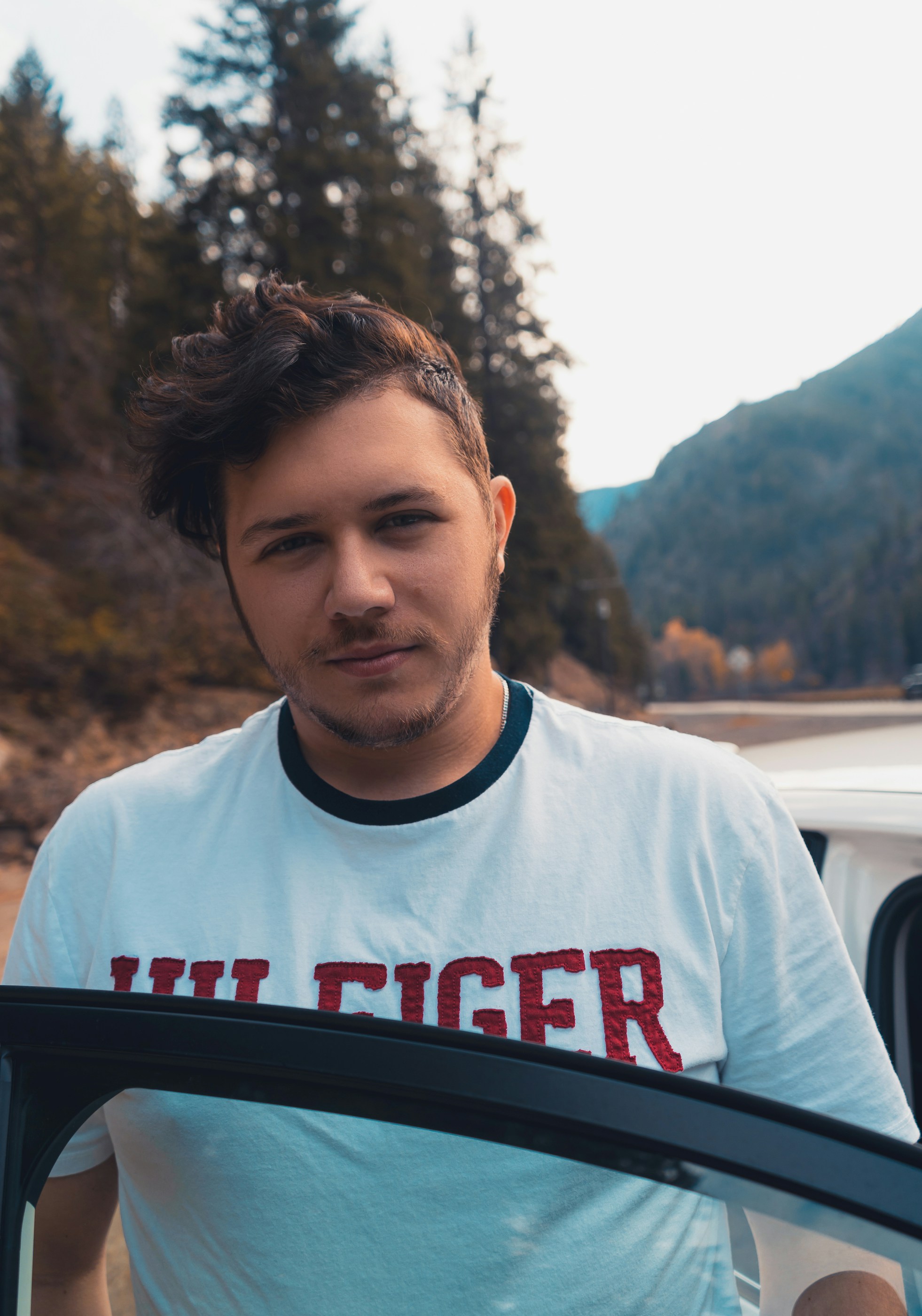A young man stands by a car door, framed by a backdrop of mountains and autumn foliage. His expression reflects a moment of contemplation amidst nature.