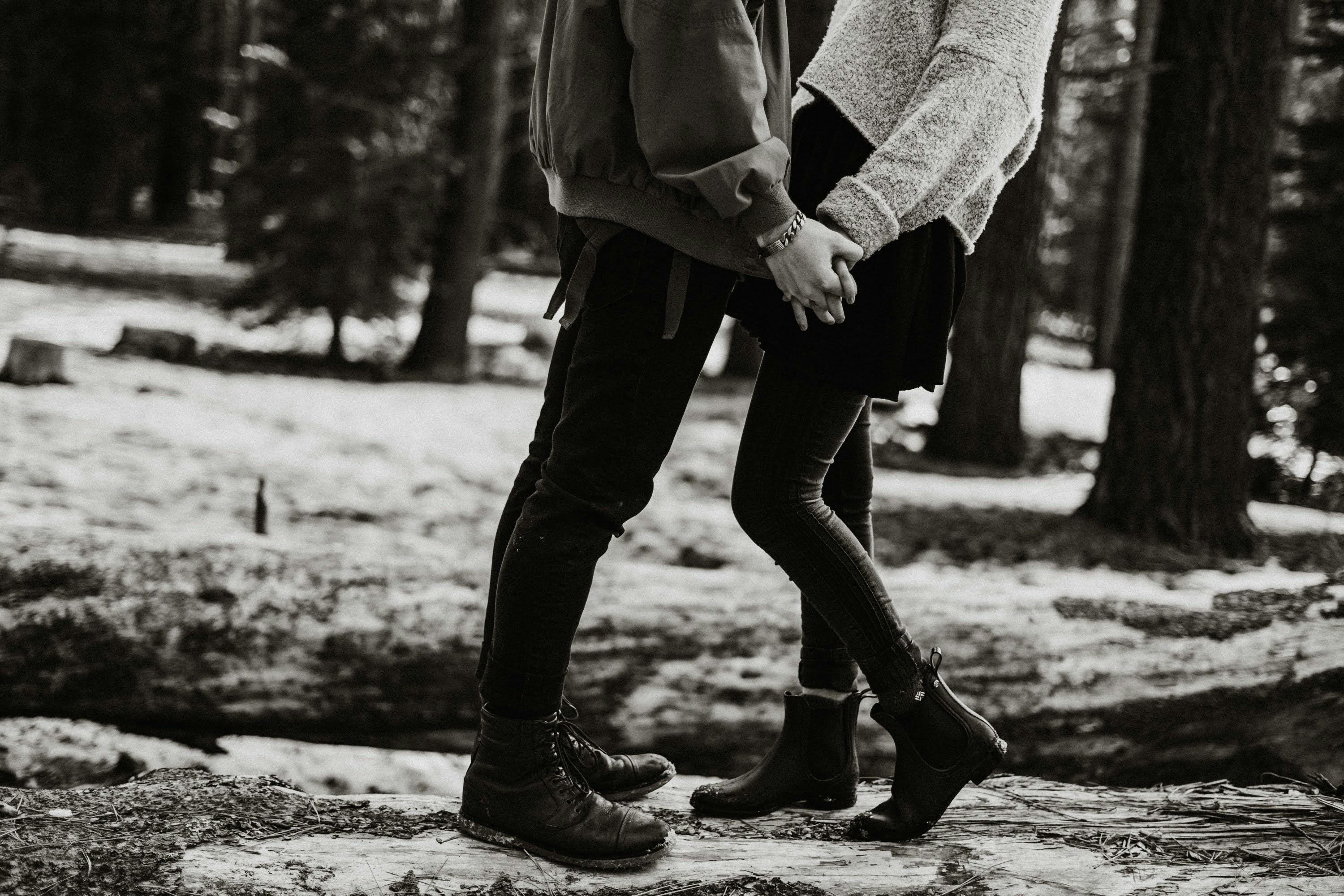 Two people holding hands in a forest, wearing boots and standing on a snowy path.