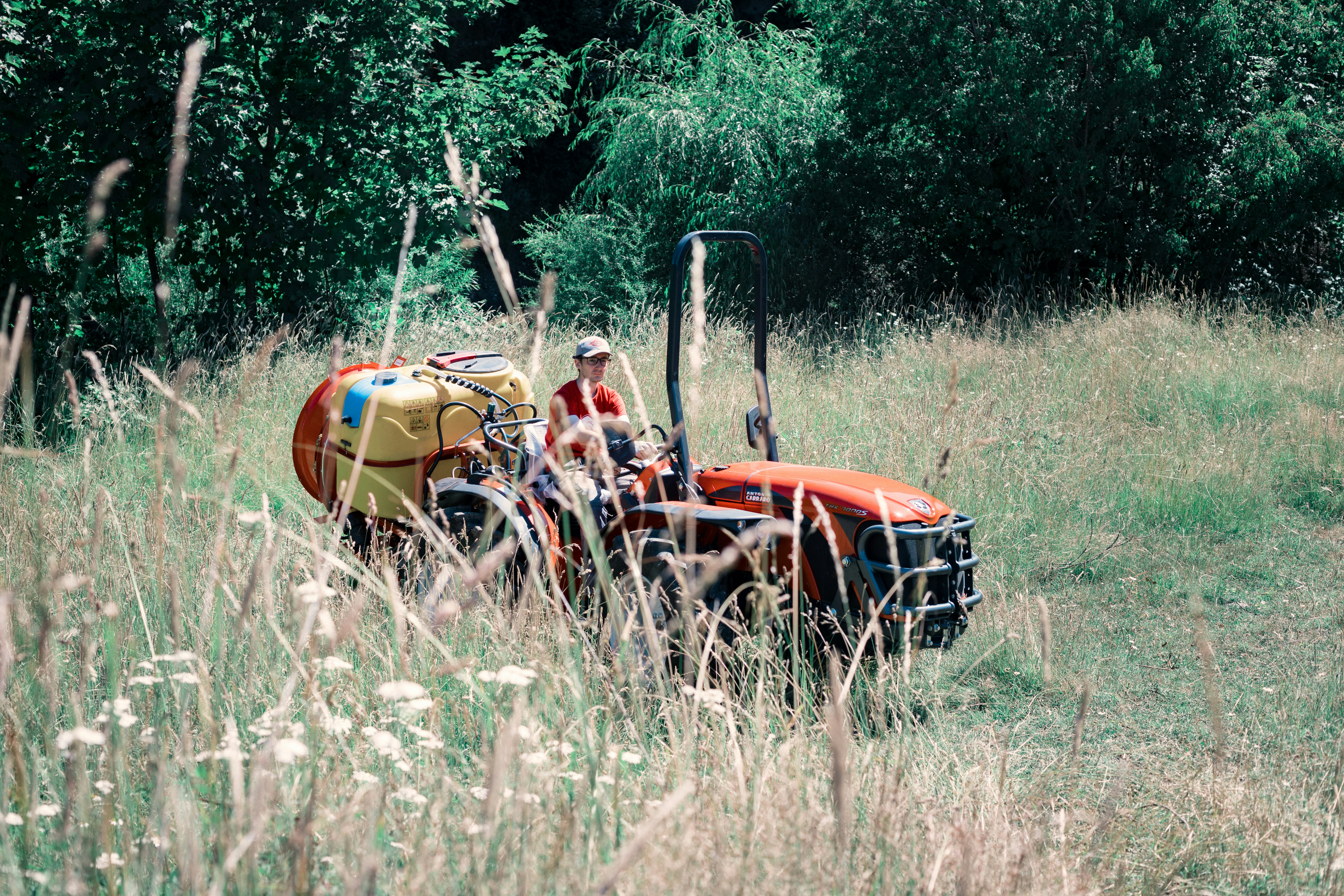A farmer operates a bright orange tractor equipped with a yellow sprayer, surrounded by tall grasses and wildflowers in a lush field.