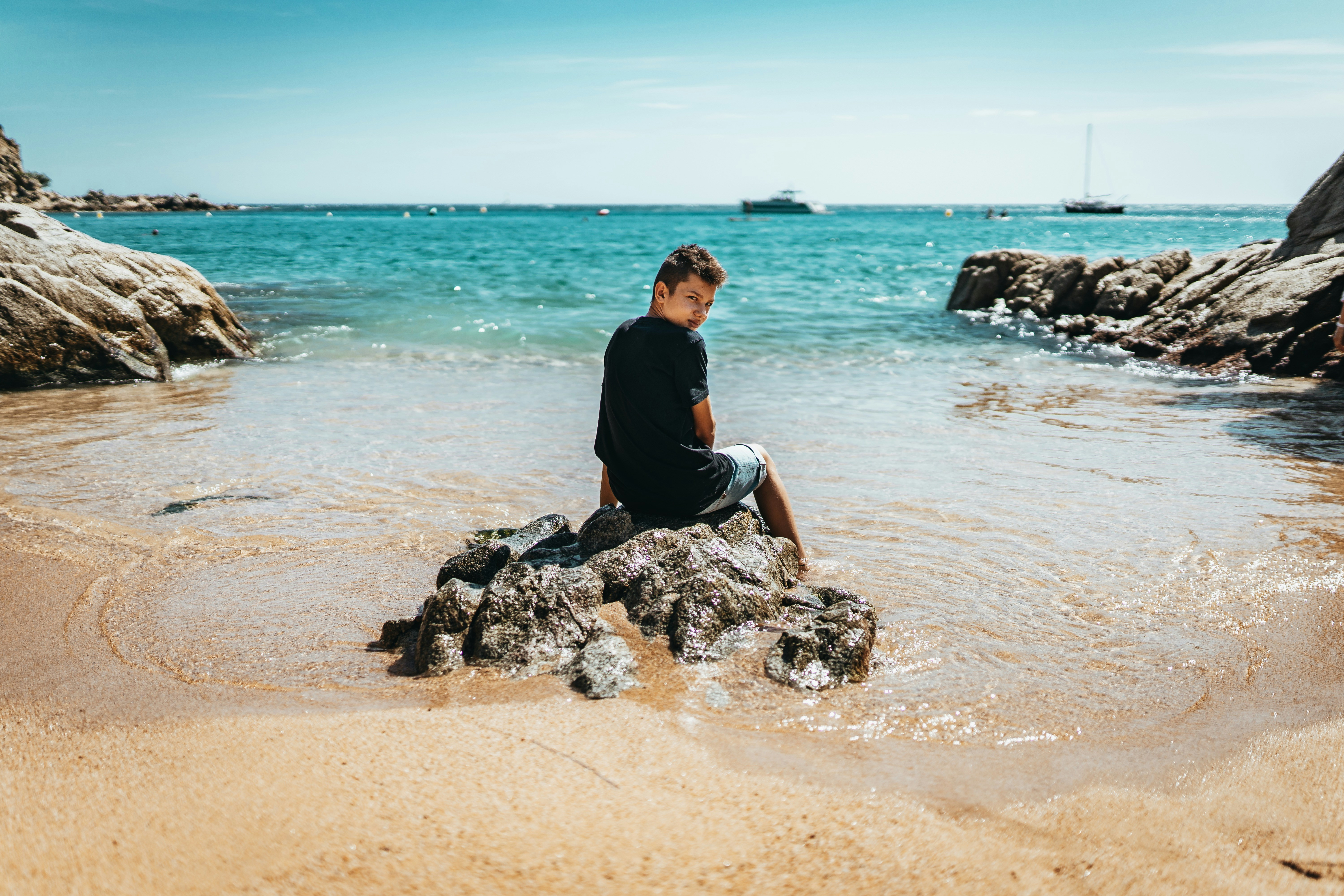 Young boy sitting on a rock in shallow waters, gazing thoughtfully at the horizon amidst a tranquil beach setting.