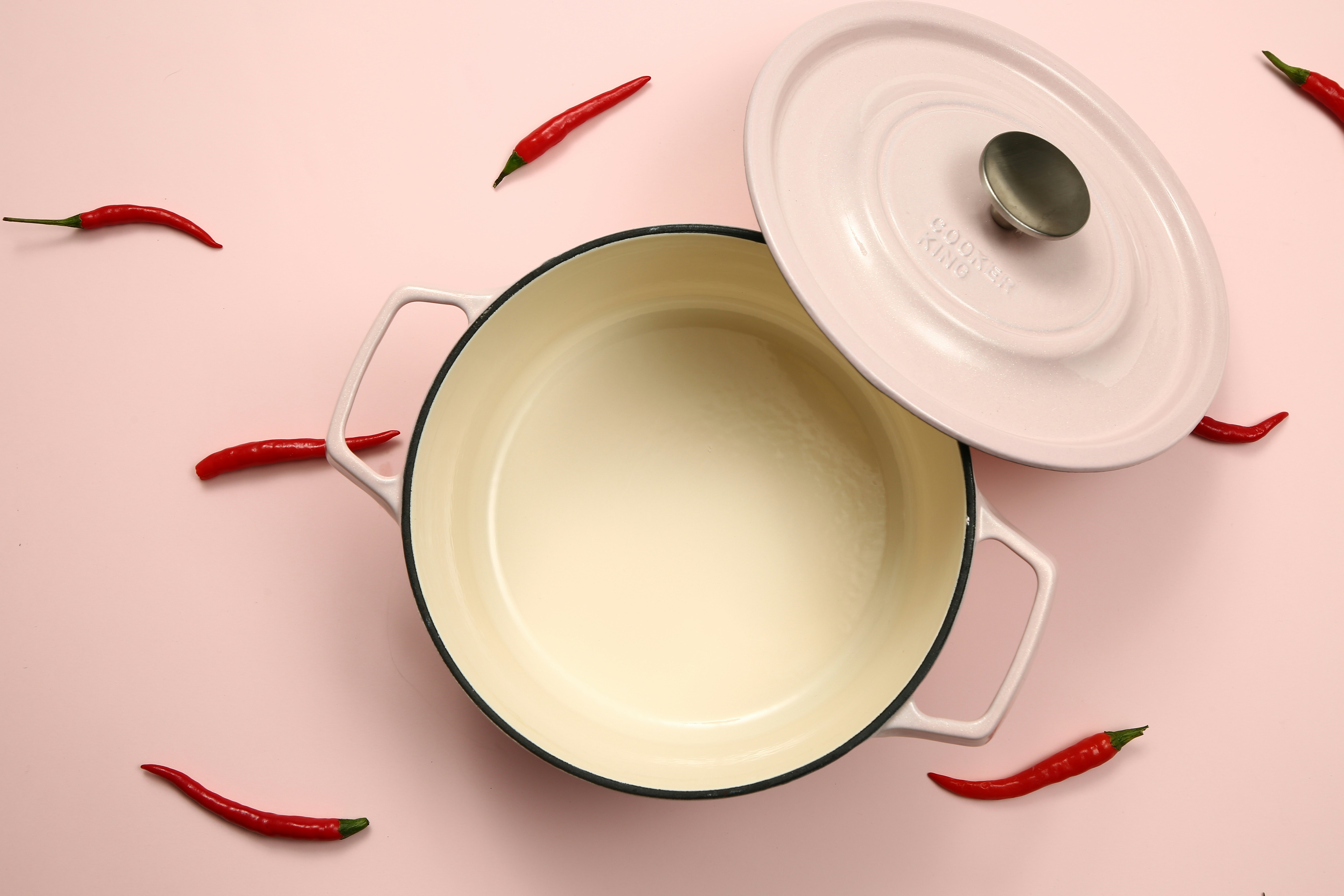 A pristine white cooking pot with an open lid, surrounded by vibrant red chili peppers on a soft pink backdrop.