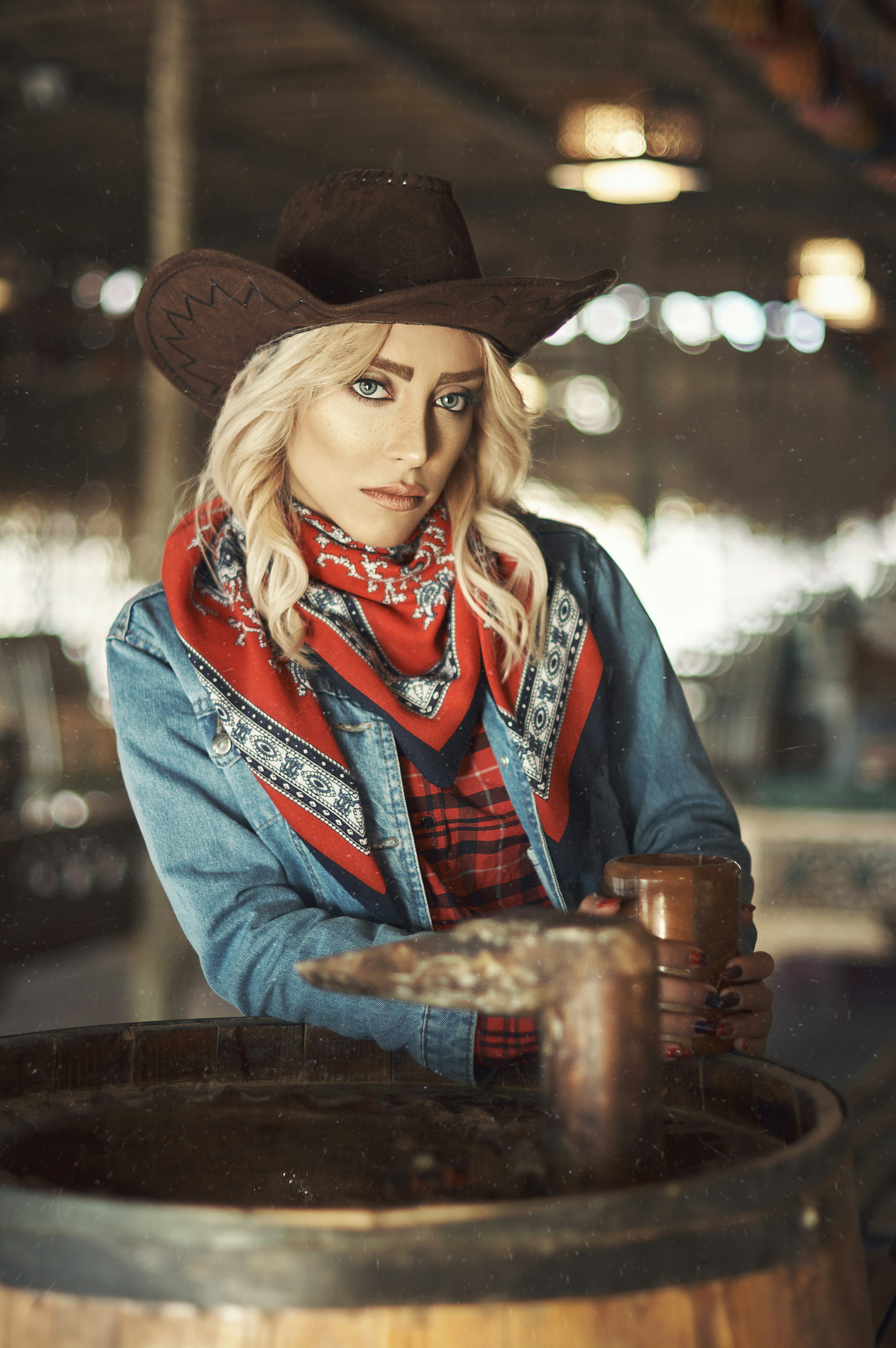A woman in a cowboy hat and bandana leans against a wooden barrel, exuding a blend of vintage and contemporary Western style.