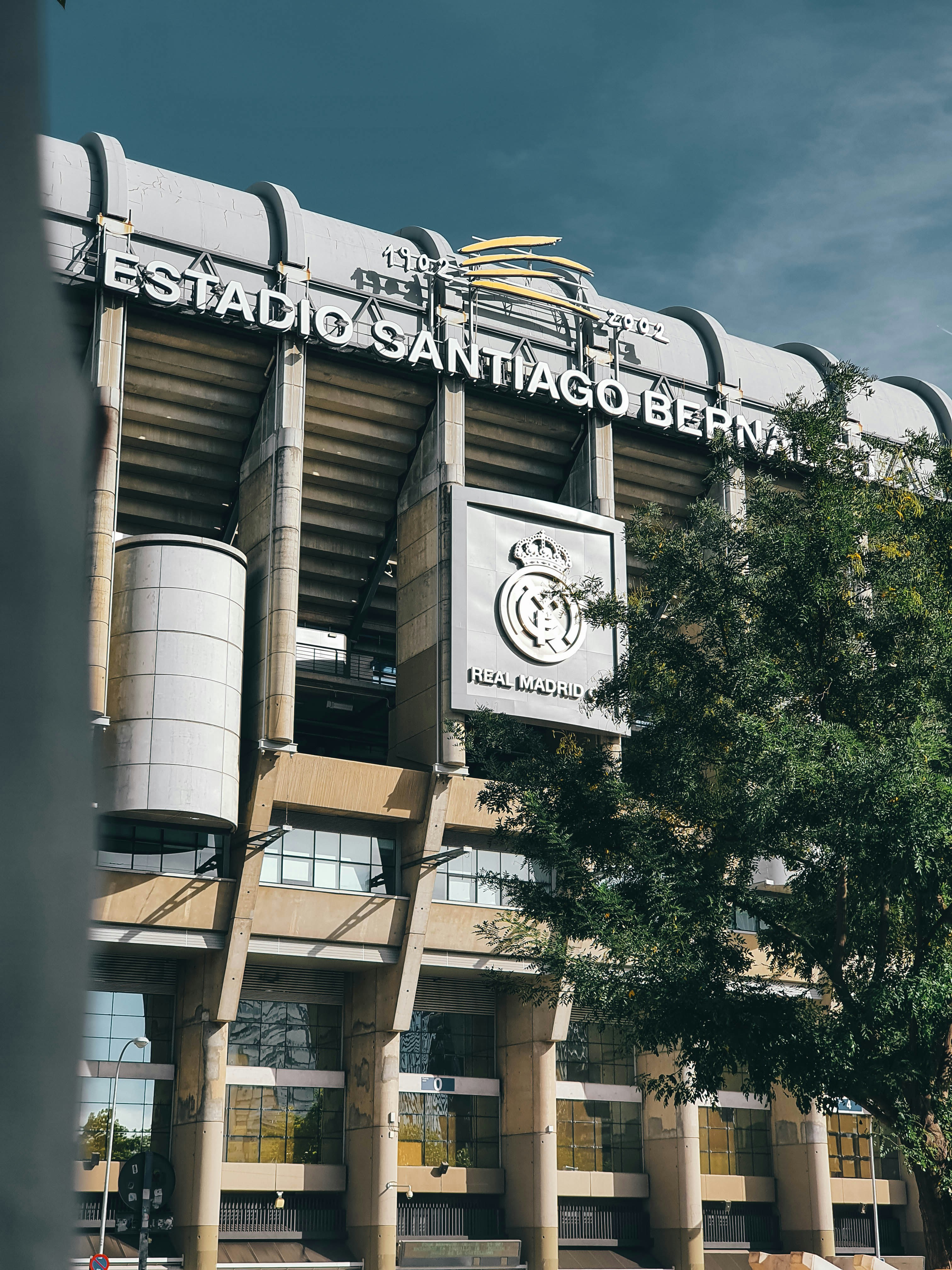 Exterior view of Estadio Santiago Bernabéu facade with the Real Madrid crest logo hanging between the concrete arches.