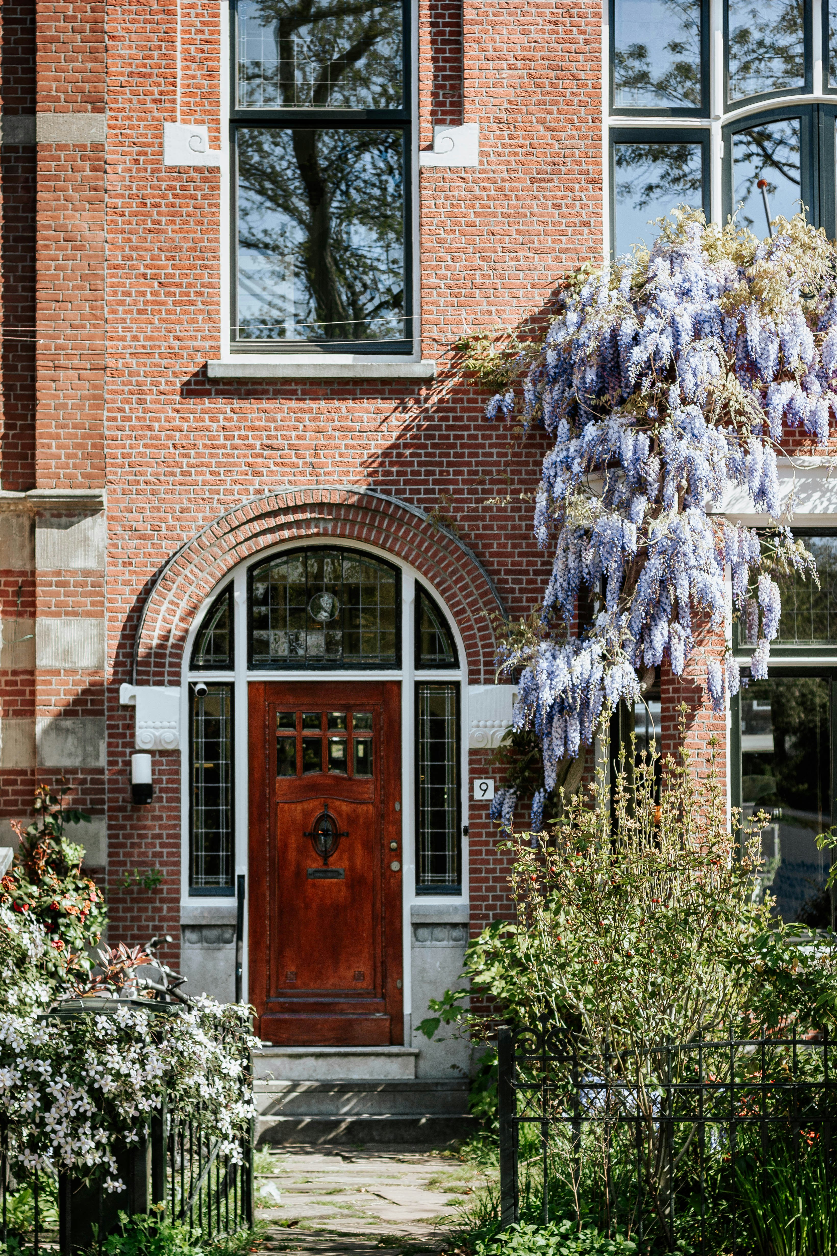 Charming brick facade adorned with cascading wisteria flowers and vibrant garden blooms. A welcoming wooden door invites exploration.