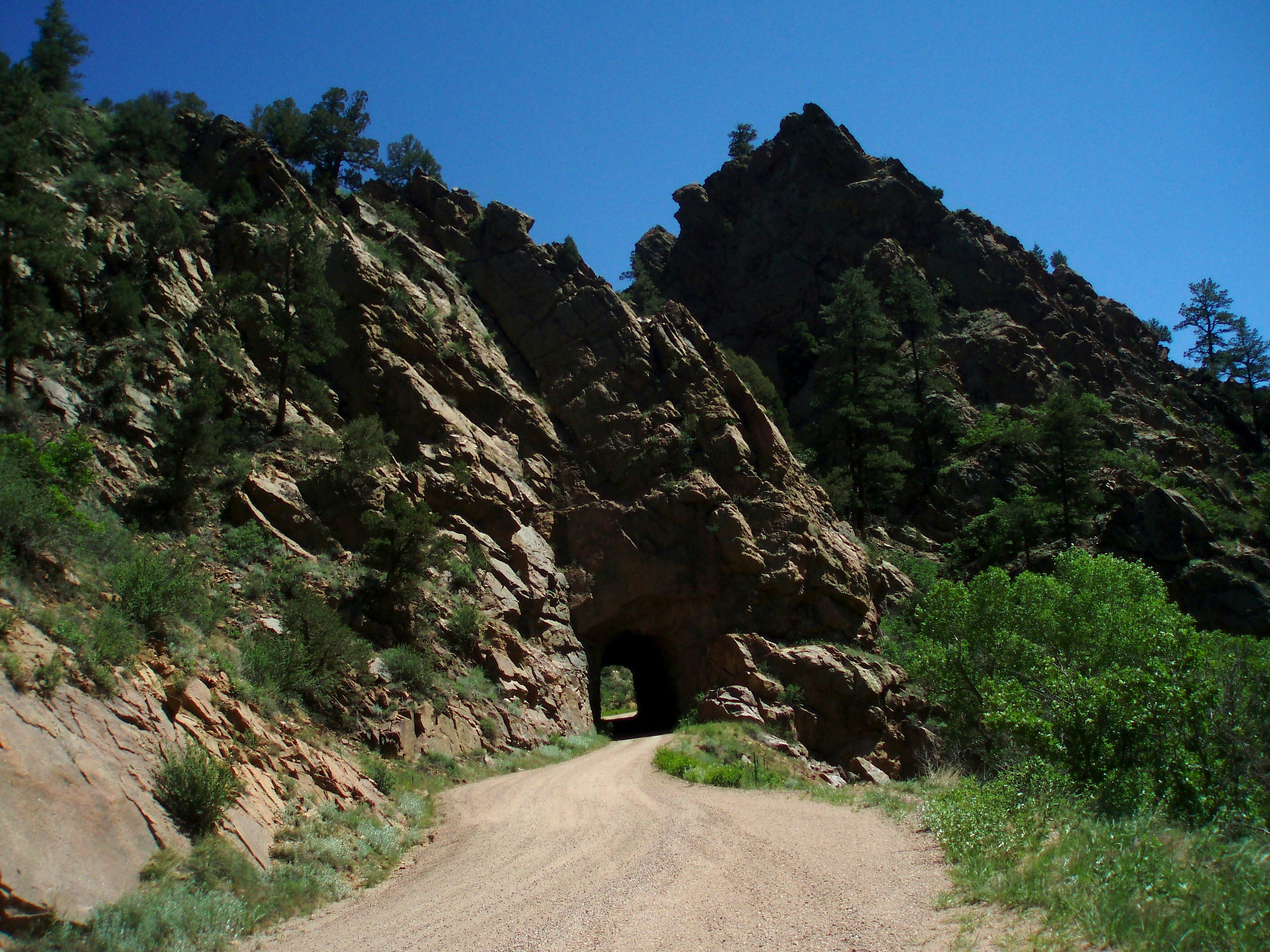 Dirt road winds through a rugged mountain pass toward a dark tunnel, framed by pines under a clear blue sky.