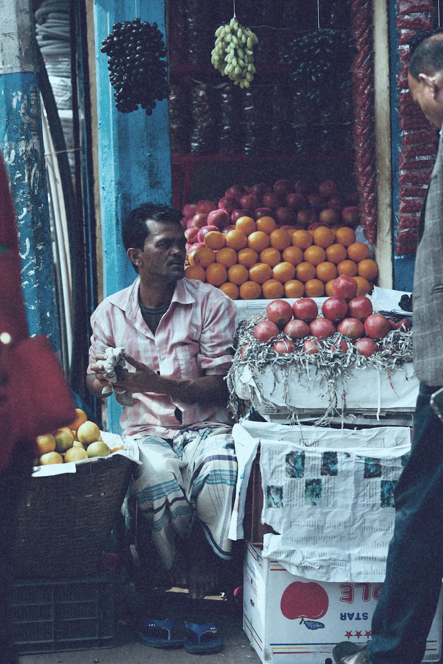 A seated fruit vendor in a pink shirt prepares produce in a narrow market stall packed with oranges, apples, and hanging grapes. A customer stands at the edge of the frame.