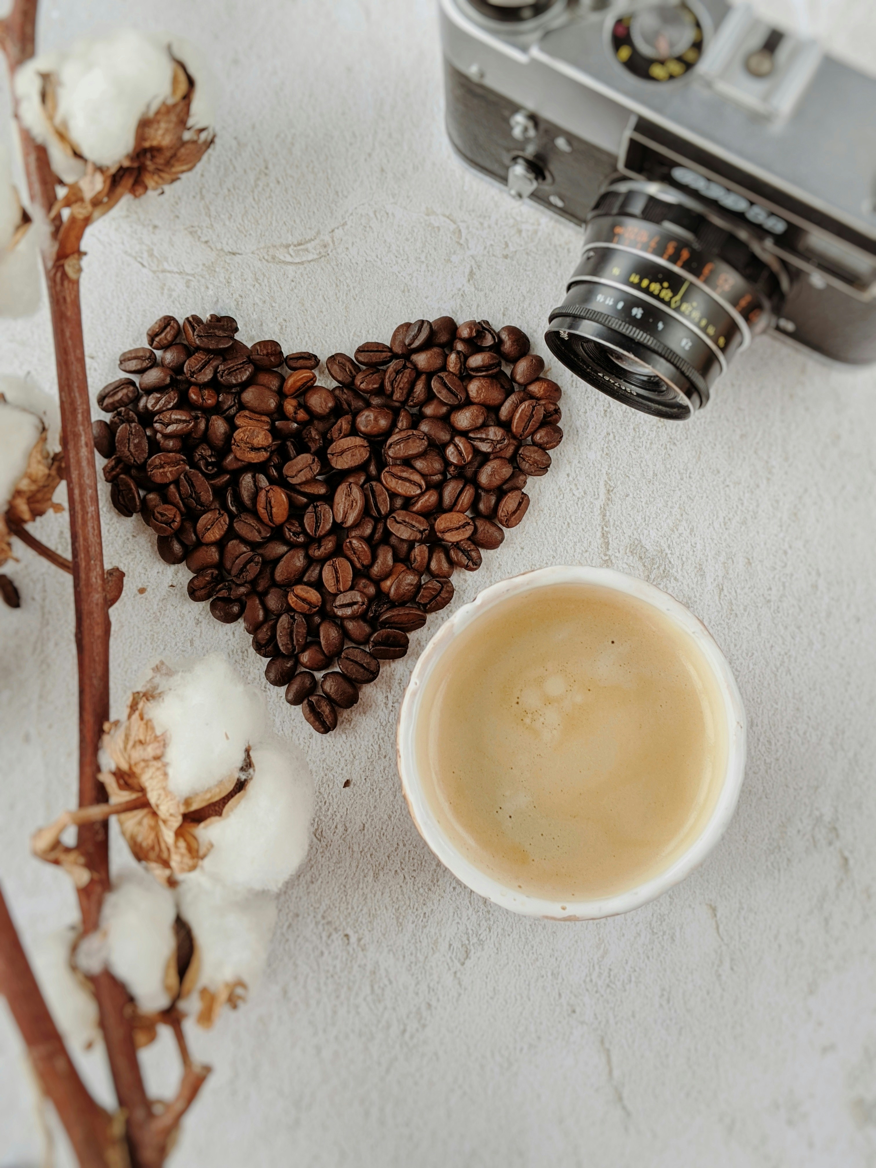 Heart-shaped arrangement of coffee beans alongside a cup of espresso and an old camera, set against a textured background. Cotton branches add a rustic touch.