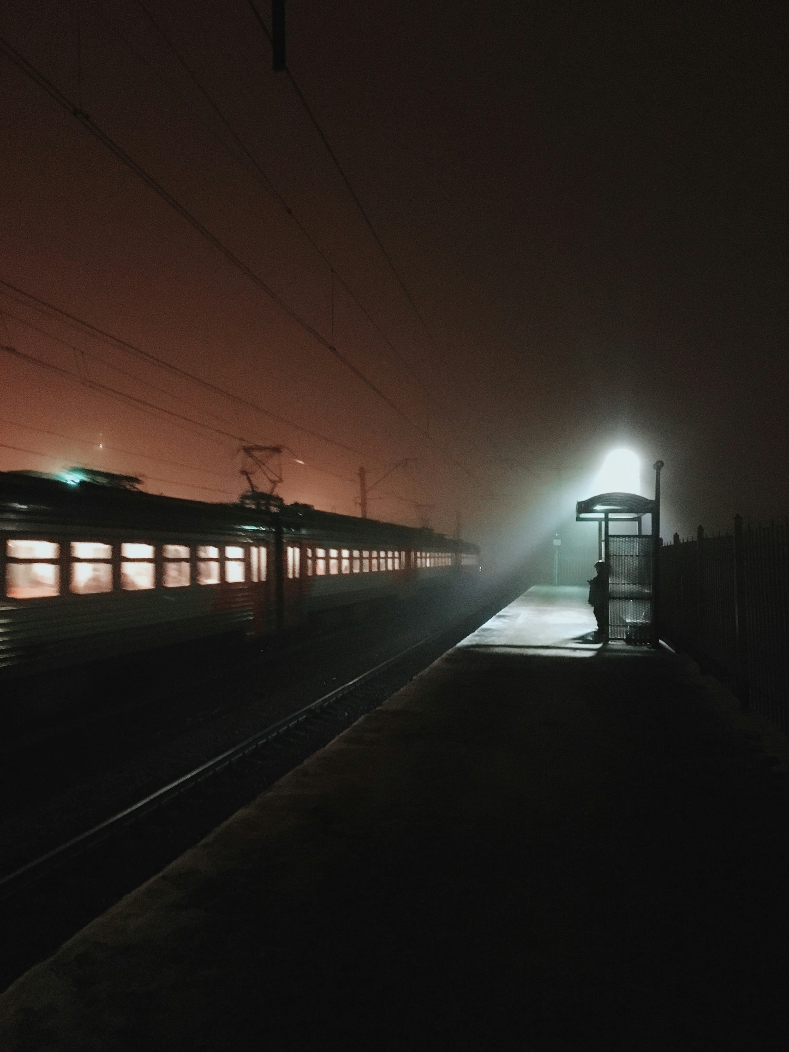 A train glides silently through a foggy night, illuminated by a distant platform light, creating an atmosphere of mystery and solitude.