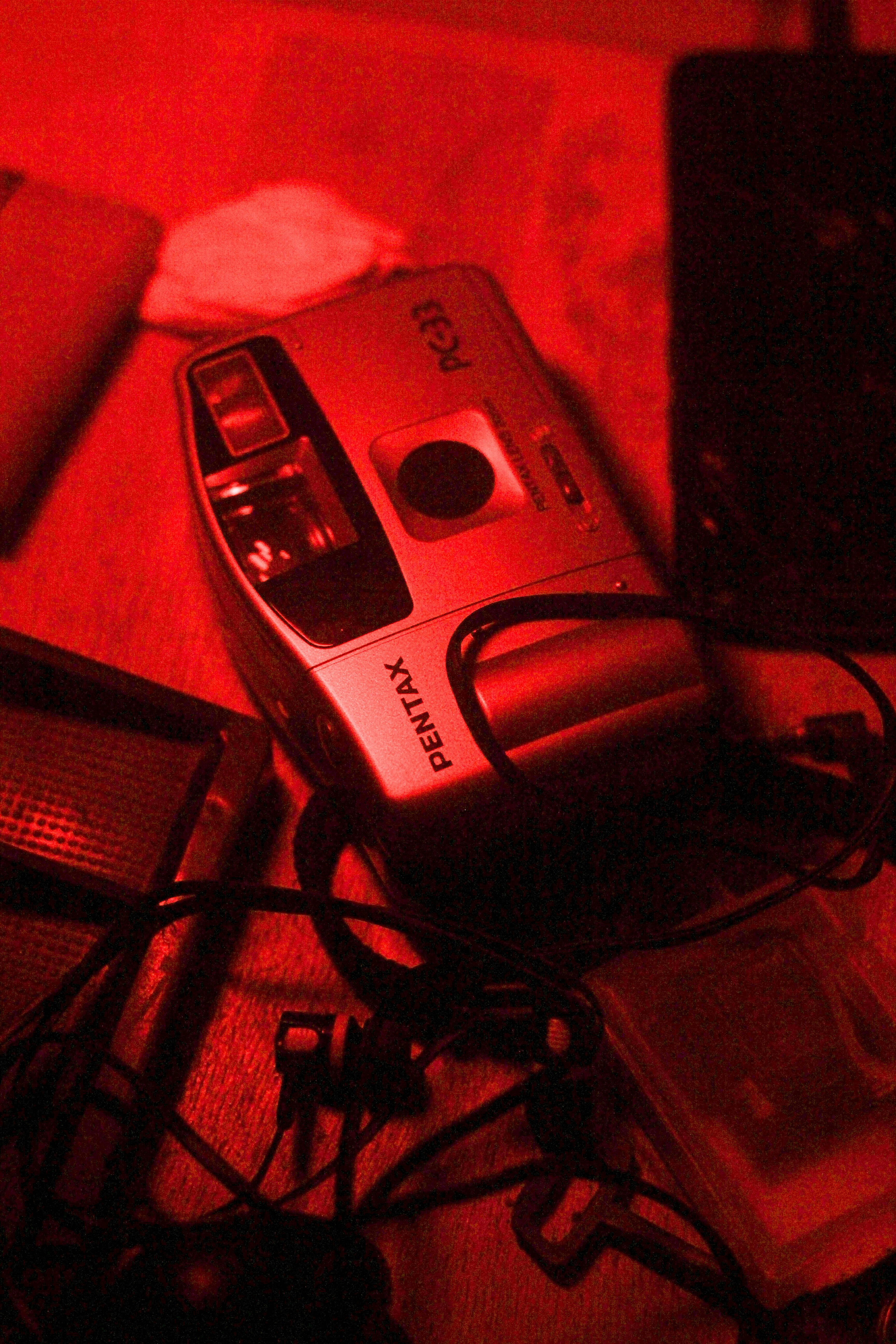 A Pentax PC-37 camera surrounded by tangled cables and objects, illuminated by a soft red light. The nostalgic atmosphere evokes memories of analog photography.