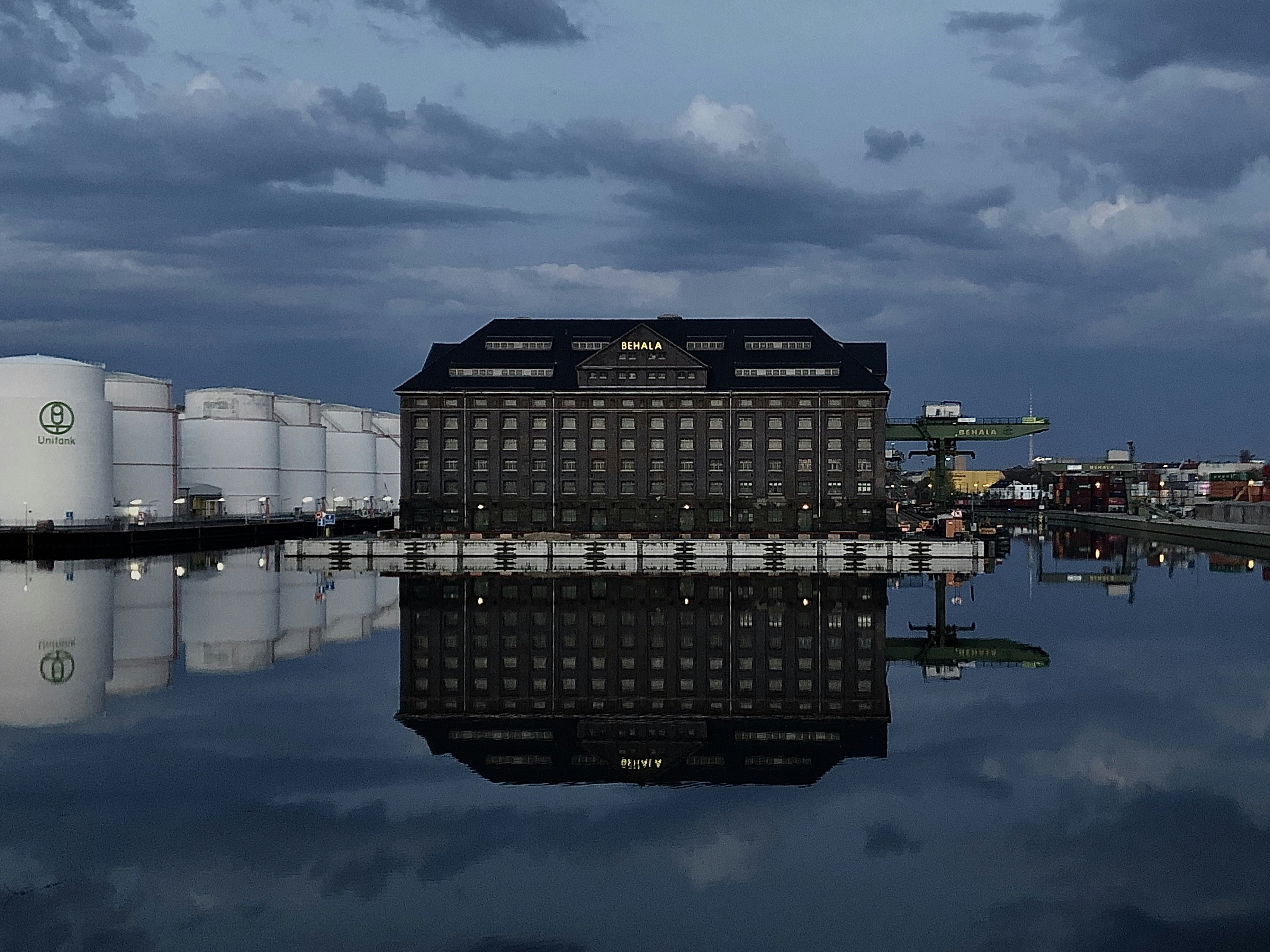 Evening view of a historic building and industrial tanks reflected in calm harbor waters under a twilight sky.