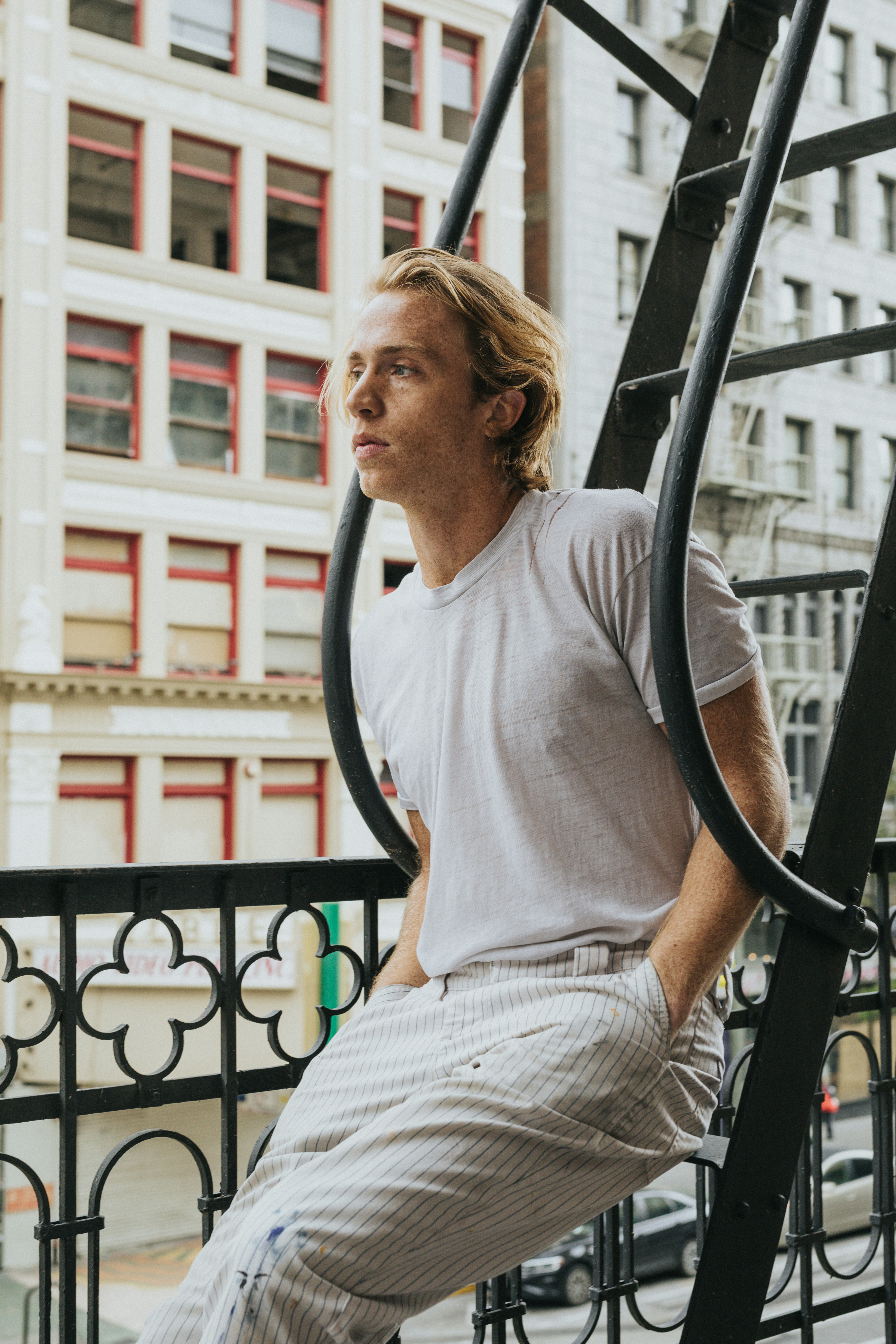Young man in a white t-shirt and striped pants leans pensively on a balcony railing, framed by a backdrop of urban architecture.