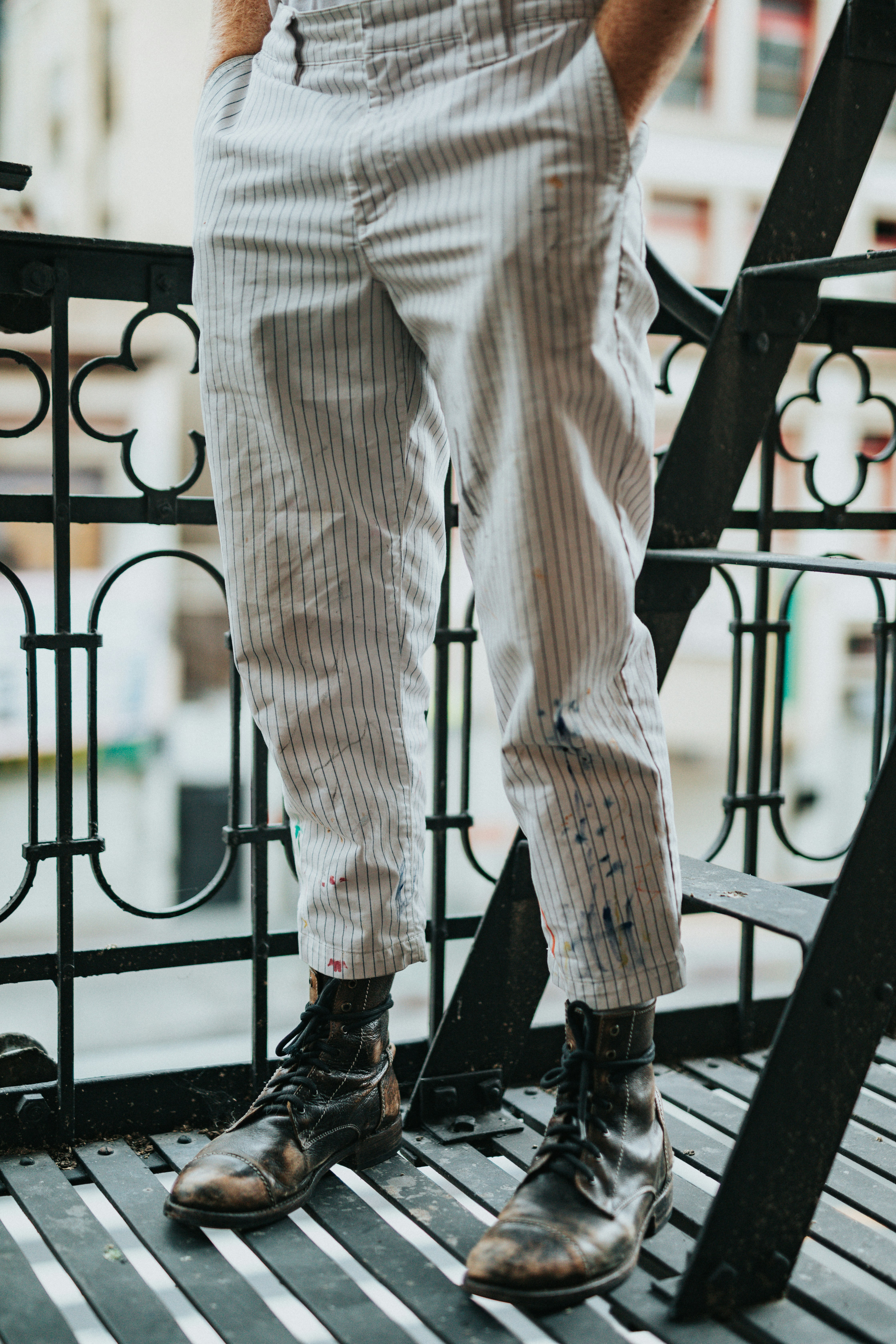 A person stands on a balcony, showcasing striped trousers paired with stylish boots, set against a blurred urban backdrop.