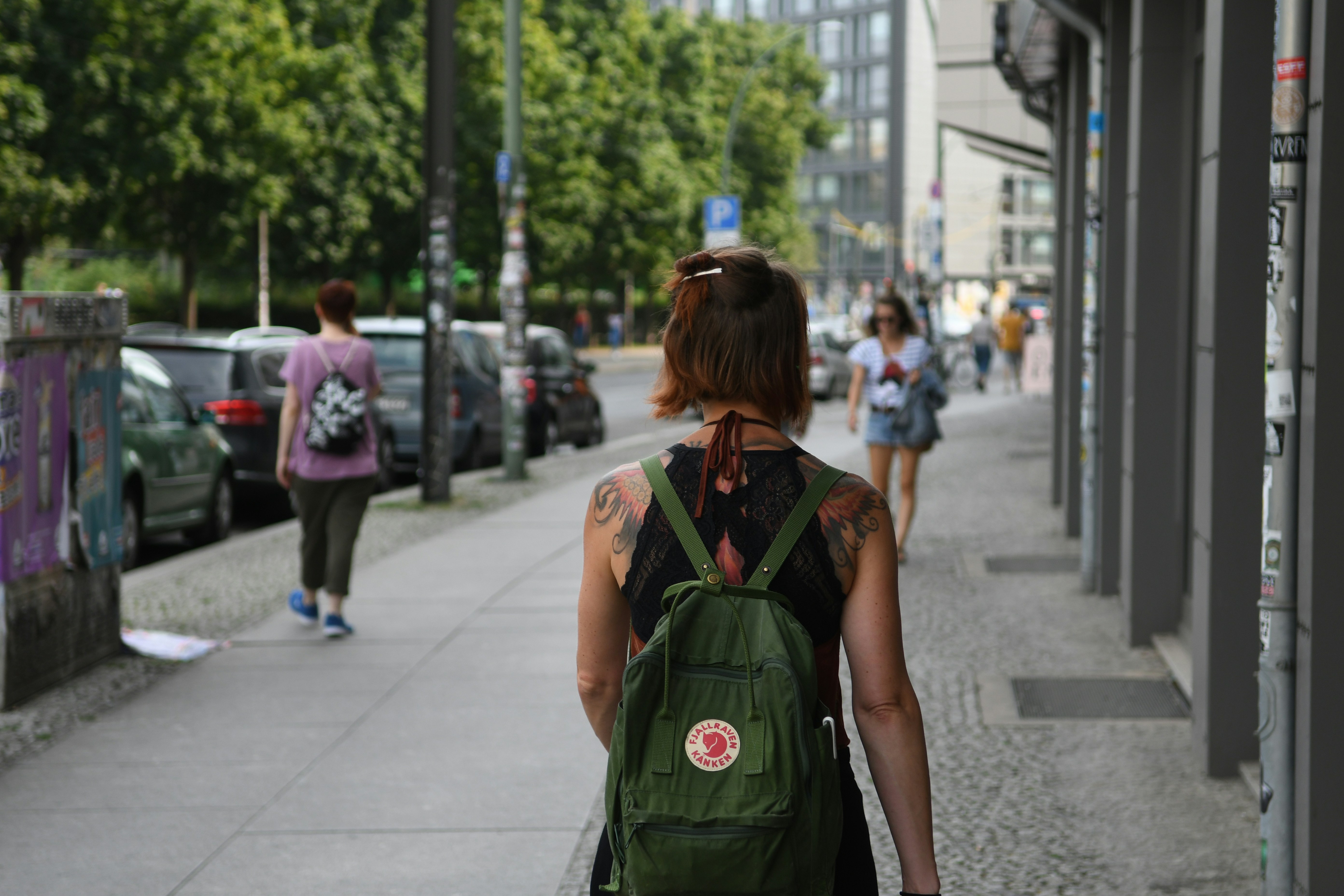 A woman with a green backpack strolls down a city sidewalk, surrounded by pedestrians and parked cars. The setting captures the essence of urban life.