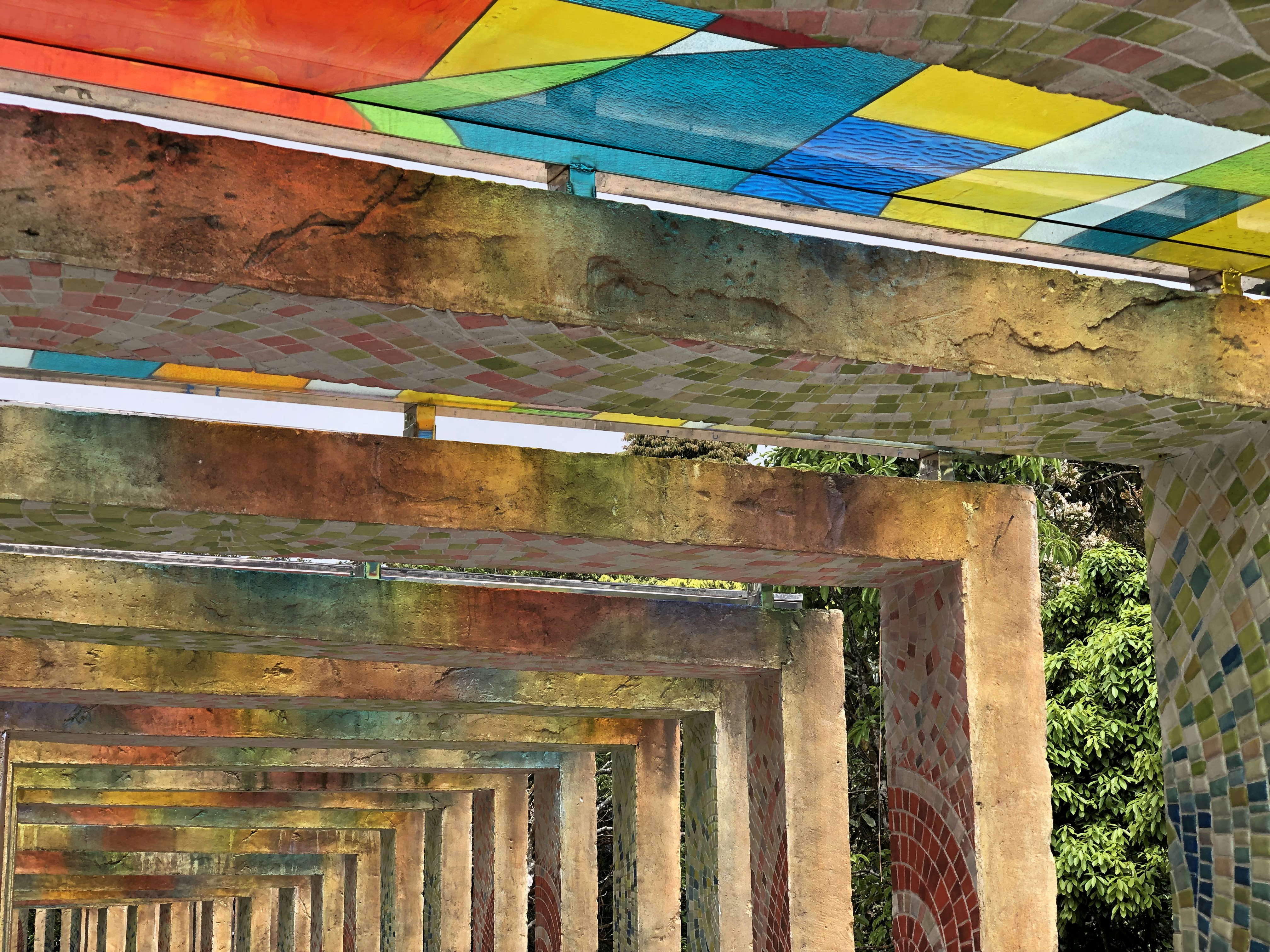 Colorful tiled patterns on concrete beams under a bridge with greenery visible in the background.