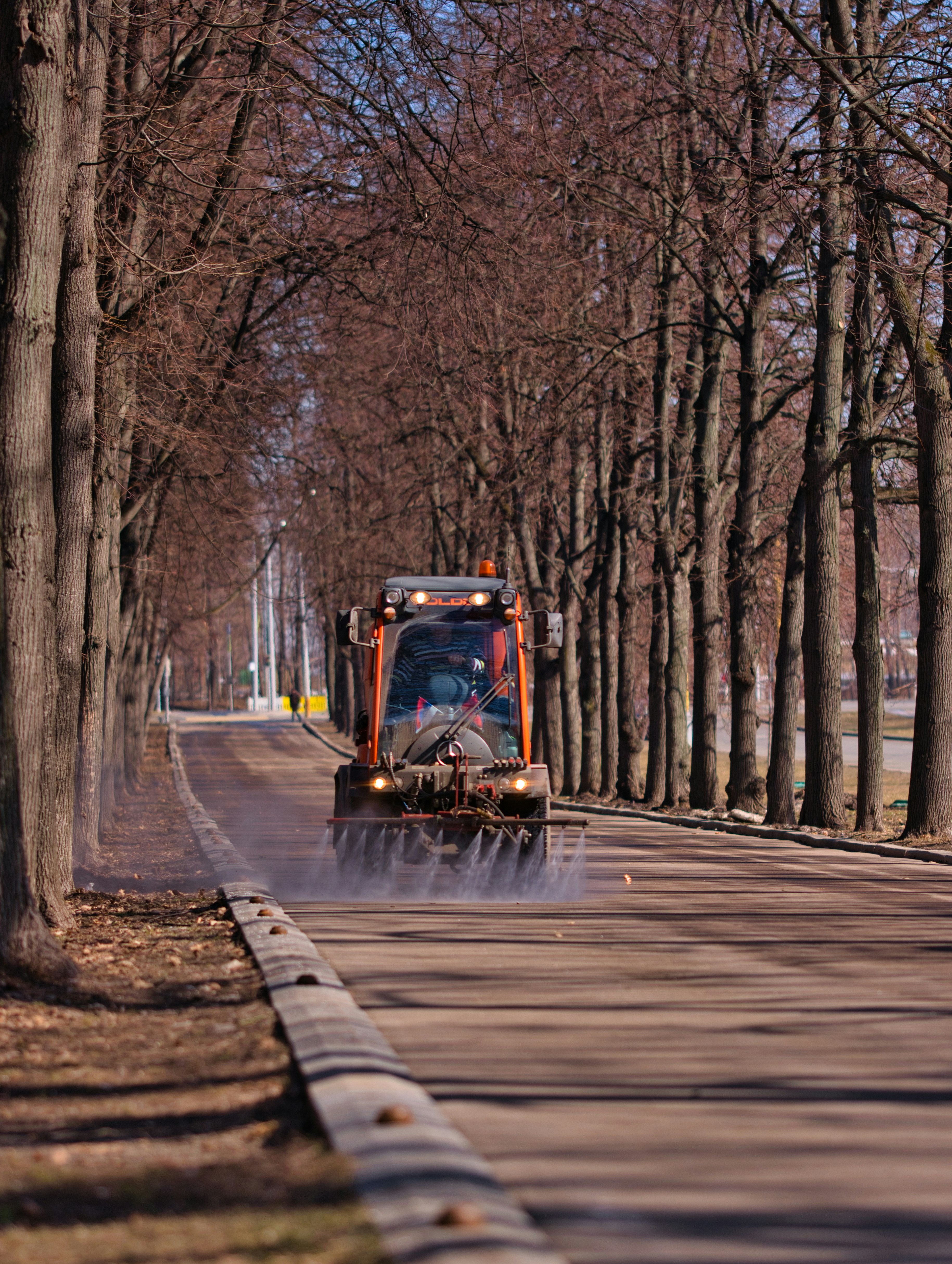 A street-sweeper moves down a quiet, tree-lined boulevard, its brushes scattering dust as it clears the lane.