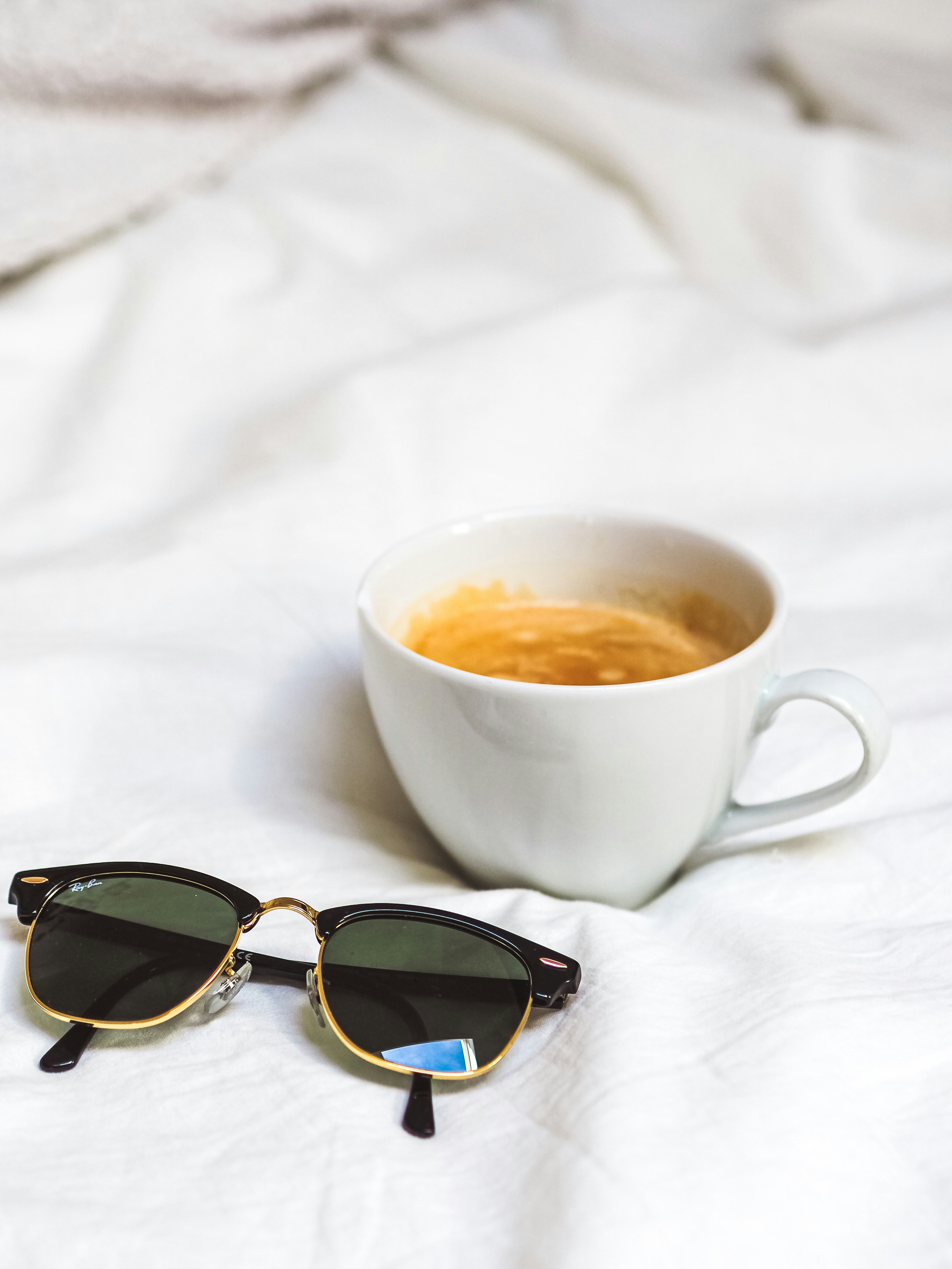 Cream cup of coffee rests on white bedsheet with a pair of sunglasses in the foreground, creating a minimalist still-life setup.