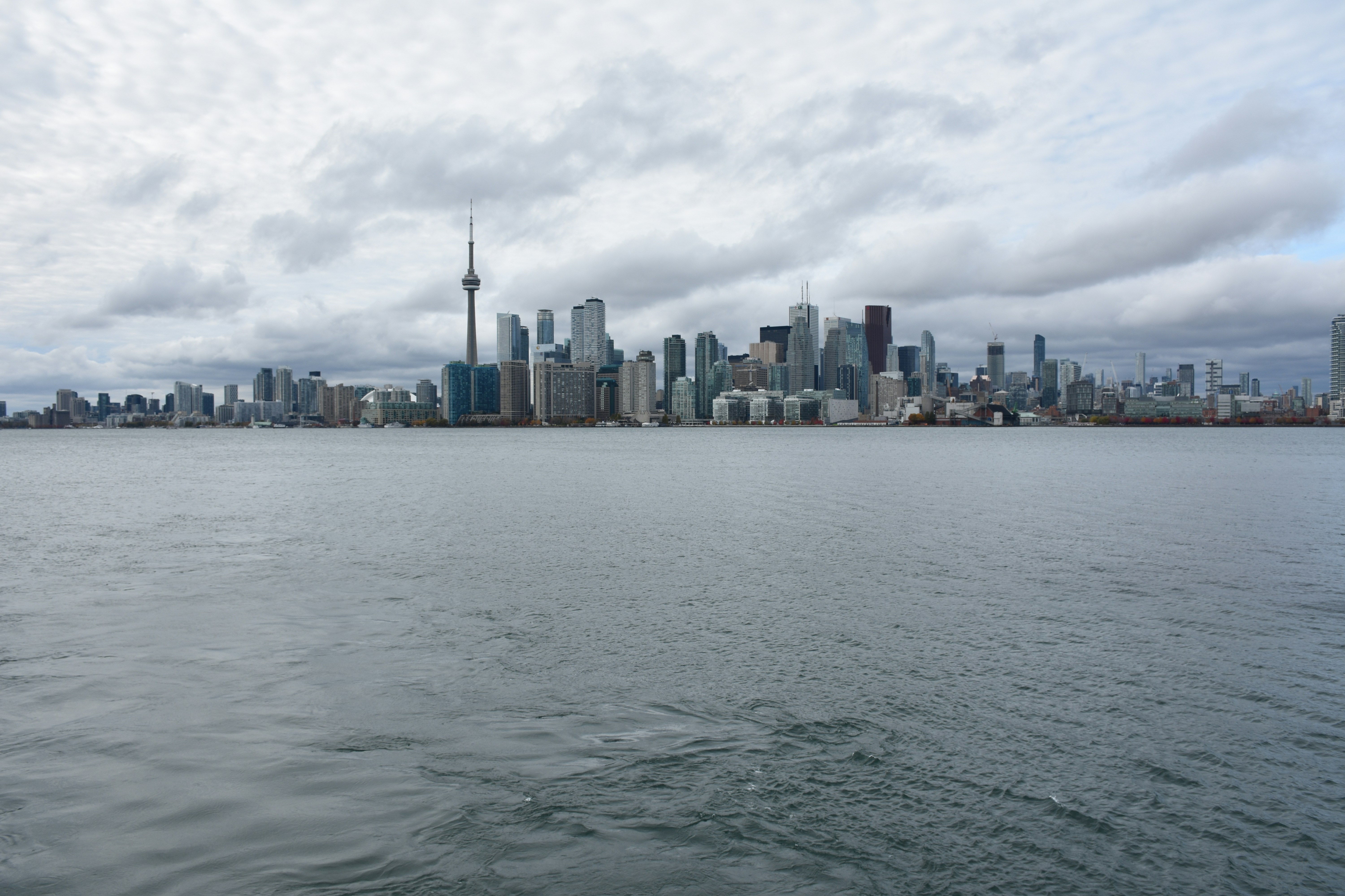 Toronto skyline with the CN Tower prominently featured, set against a backdrop of overcast skies and calm waters.