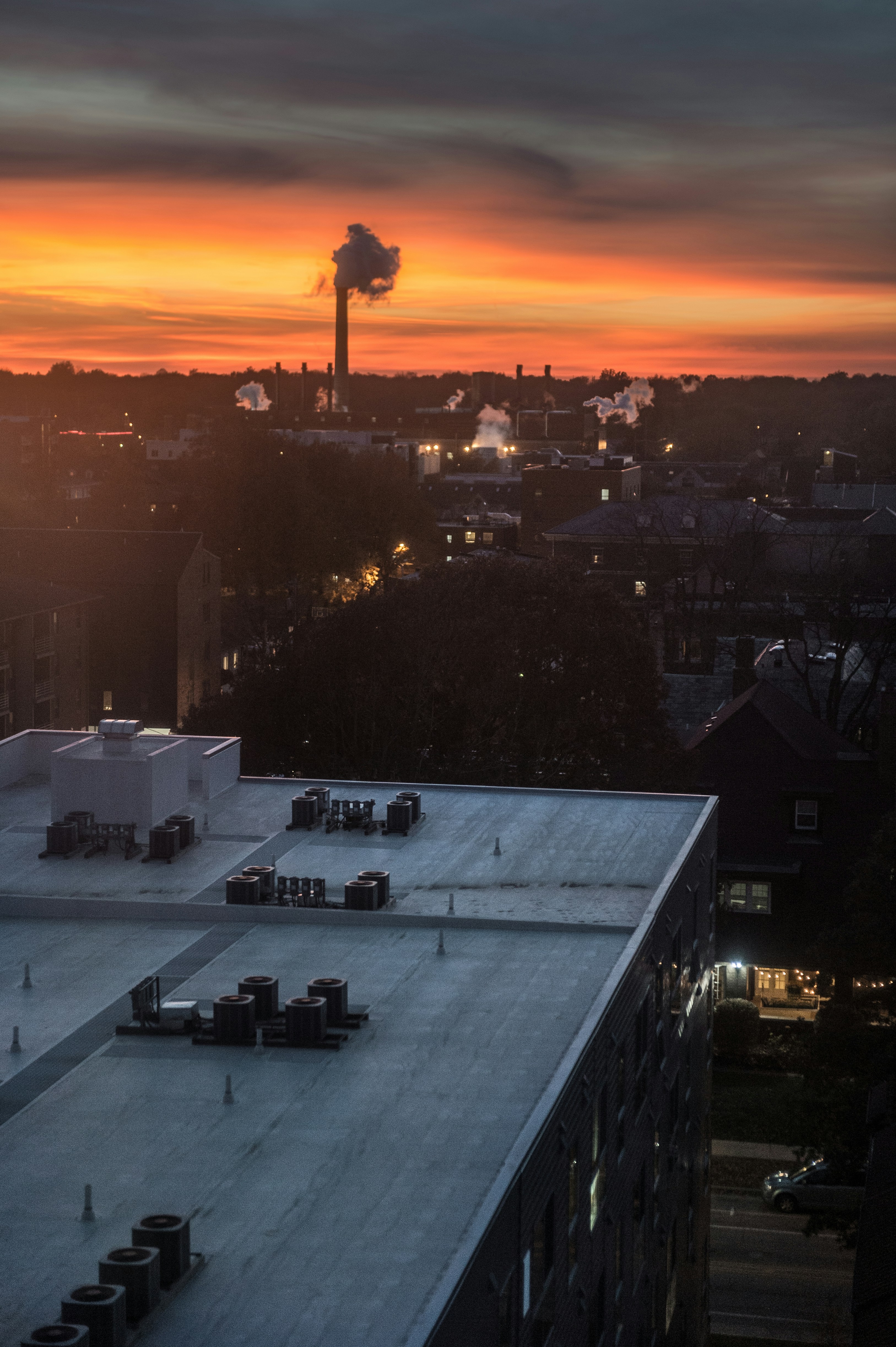 Smoke rises from factory chimneys against a vibrant sunset, illuminating the urban skyline. Rooftops and buildings create a layered perspective of the city.