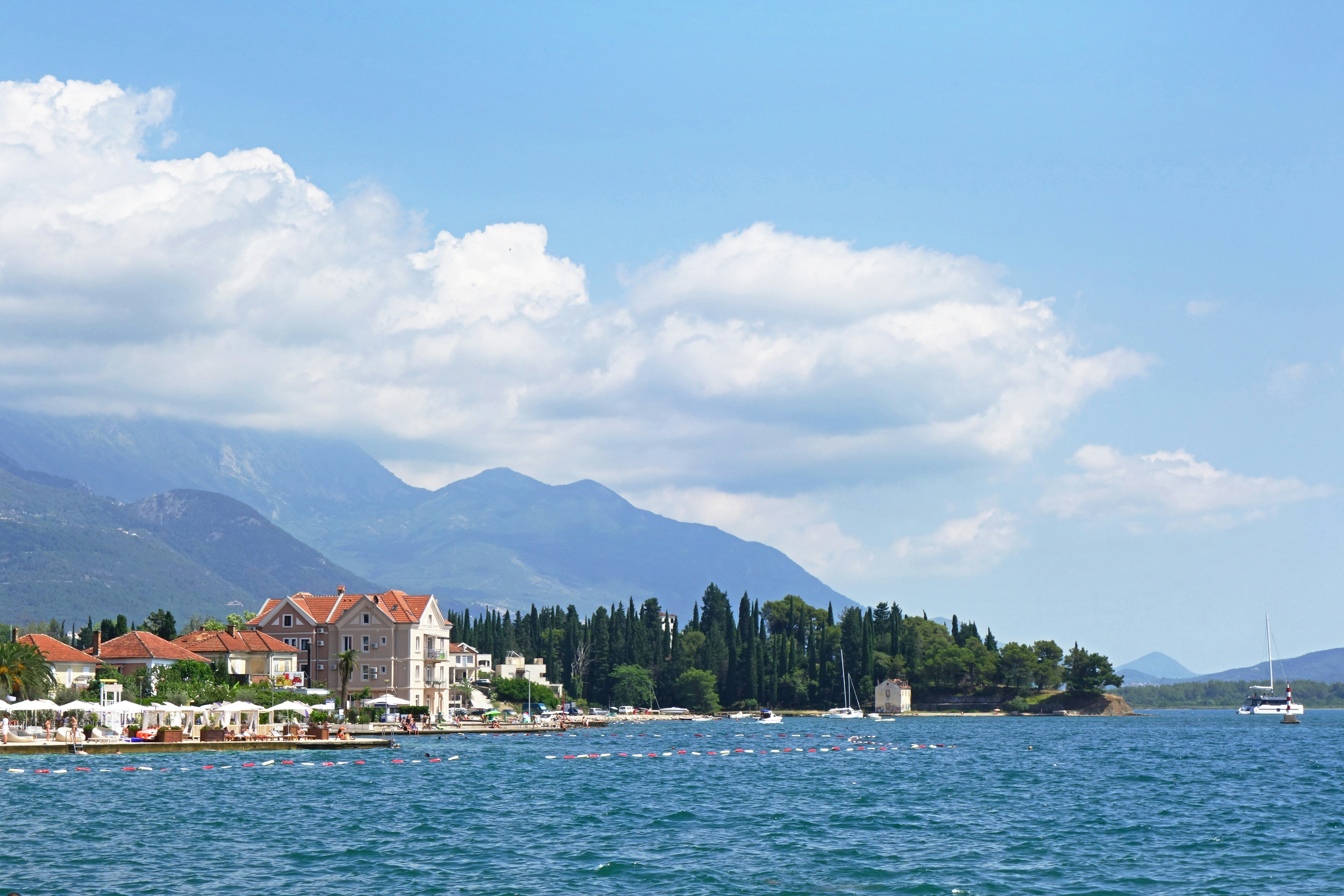 Scenic waterfront view showcasing charming coastal architecture against a backdrop of mountains and lush greenery. A sailboat glides peacefully in the distance.