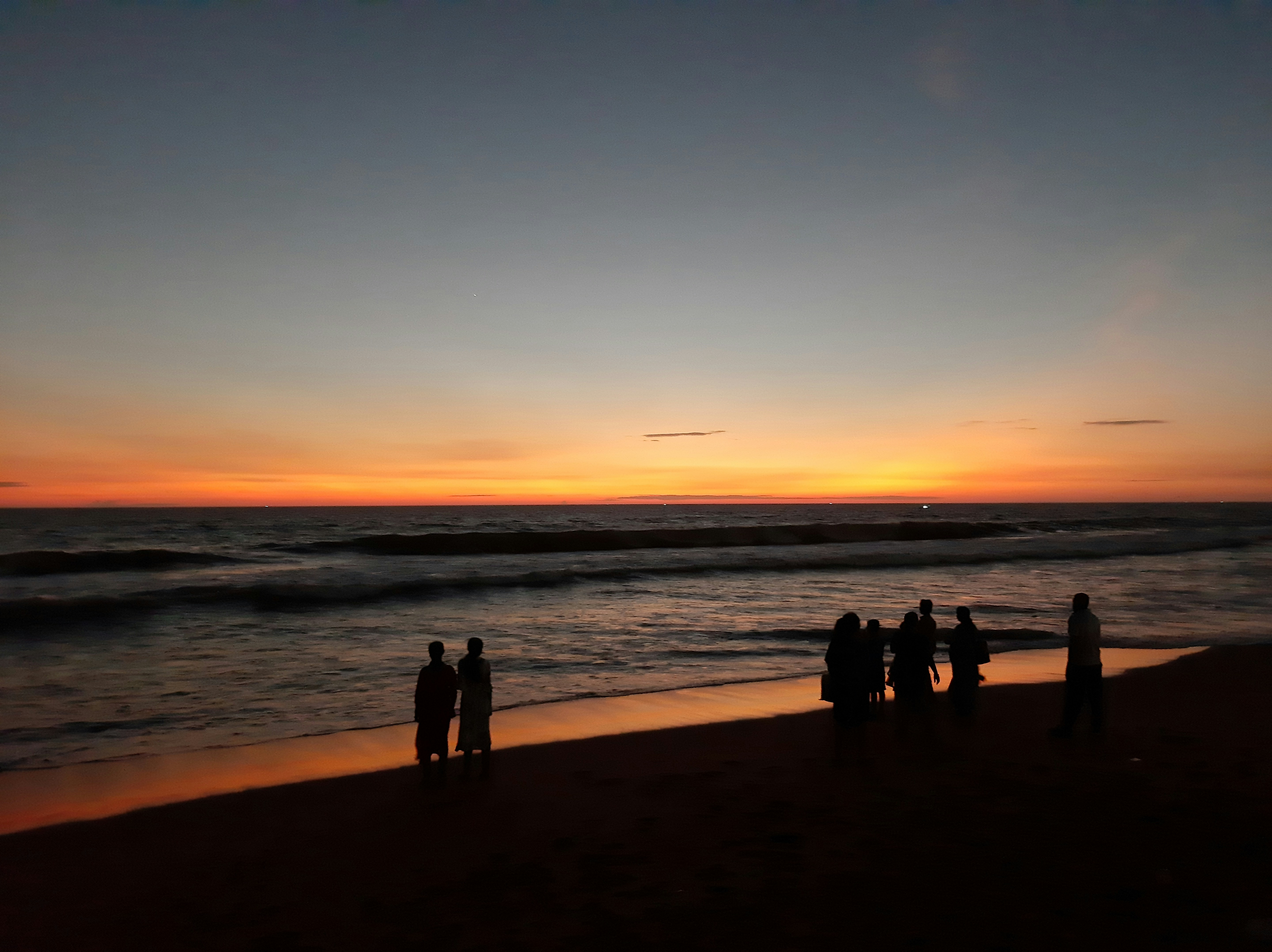 Silhouetted figures gather on the beach as the sun sets, casting a warm glow across the horizon. The tranquil waves lap gently at the shore.