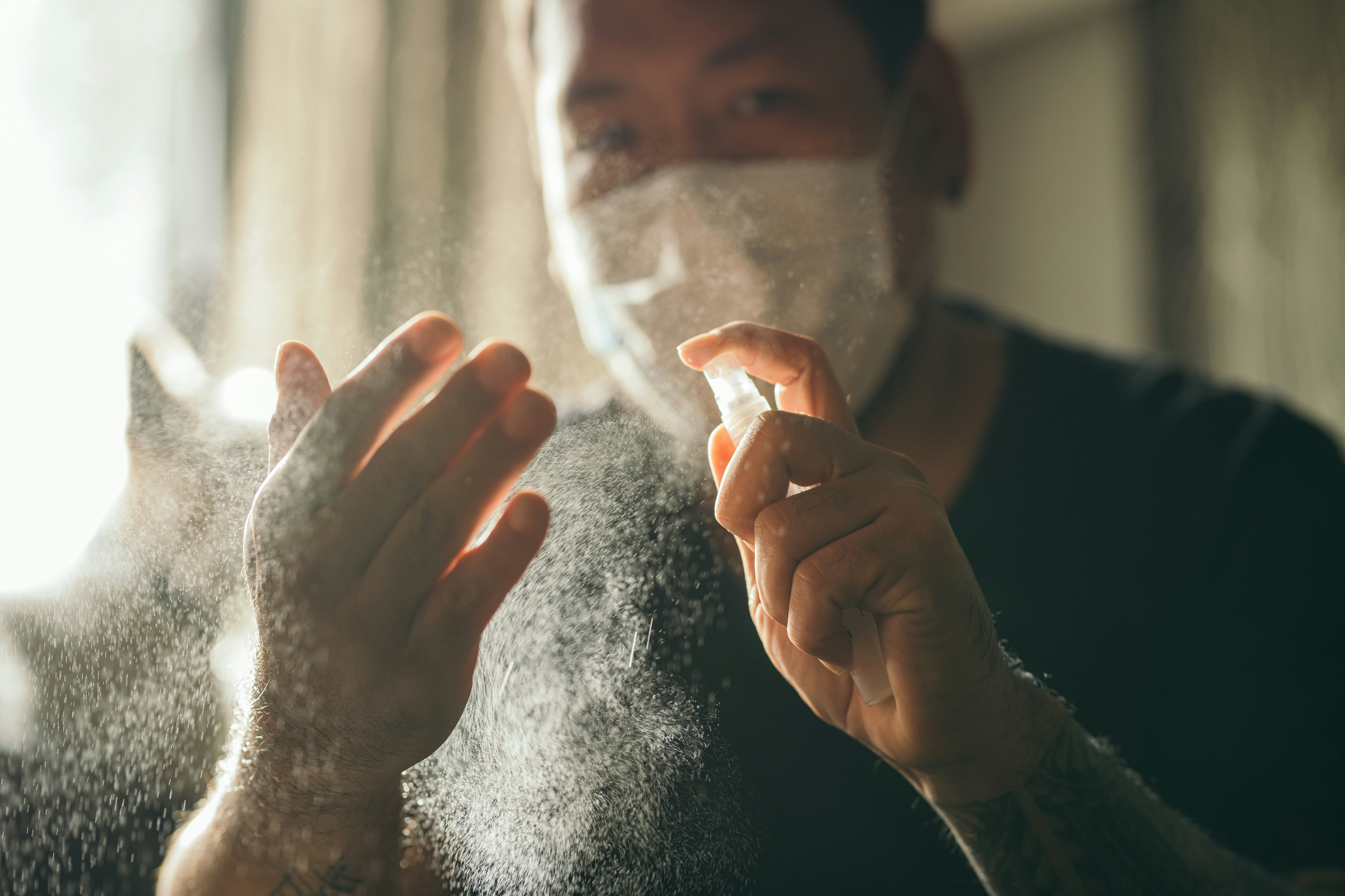 A person applies a fine mist to their hands, emphasizing personal care and hygiene. The soft light highlights the delicate particles in the air.