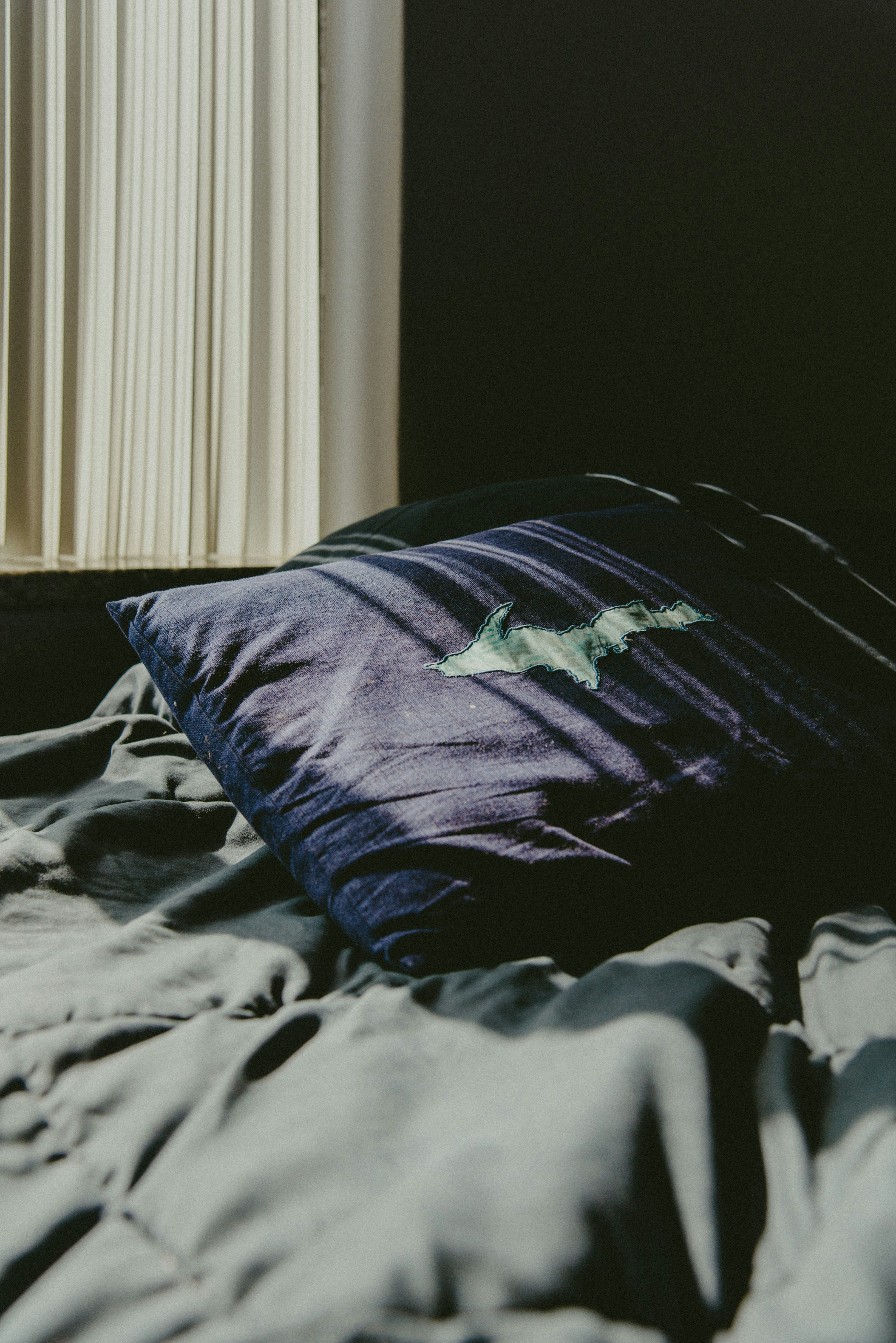 A plush navy pillow with a delicate shark embroidery rests on a rumpled bedspread, illuminated by soft, filtered sunlight from nearby blinds.