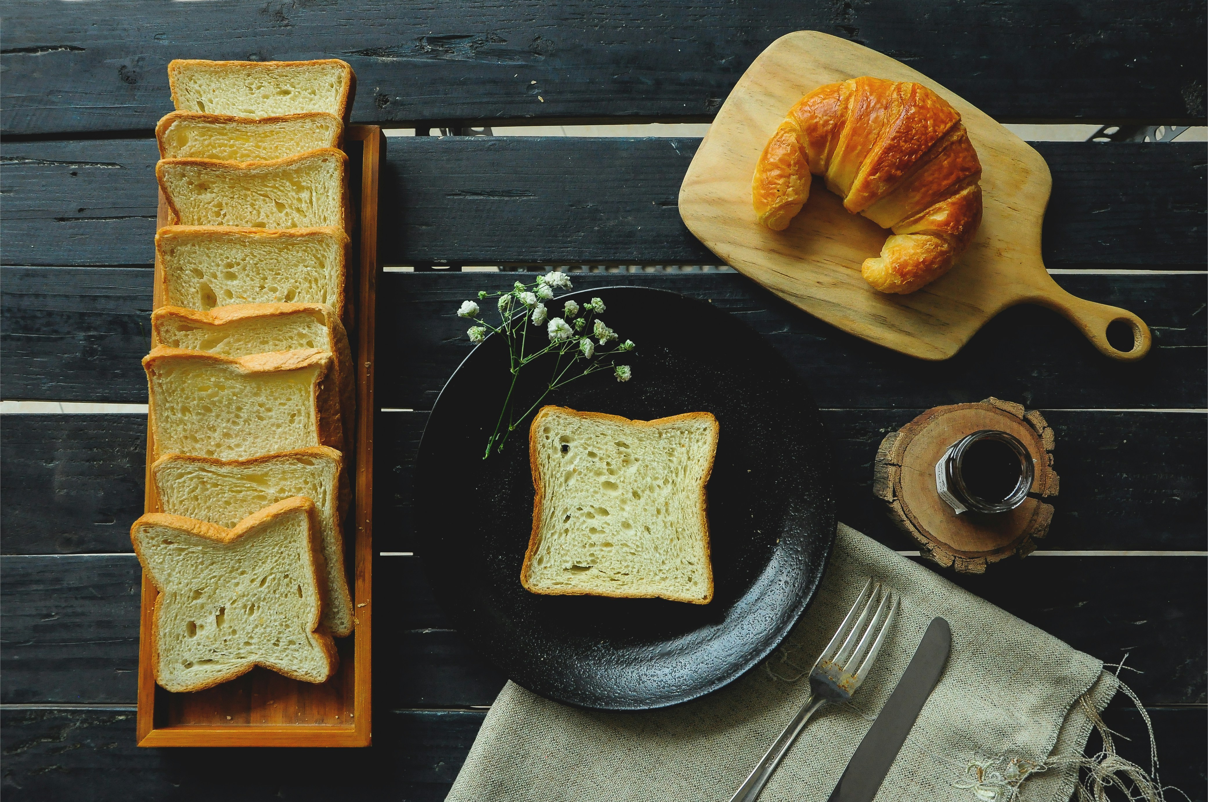 Sliced bread arranged on a wooden board alongside a croissant on a serving platter, with delicate flowers and a jar of jam enhancing the breakfast scene.