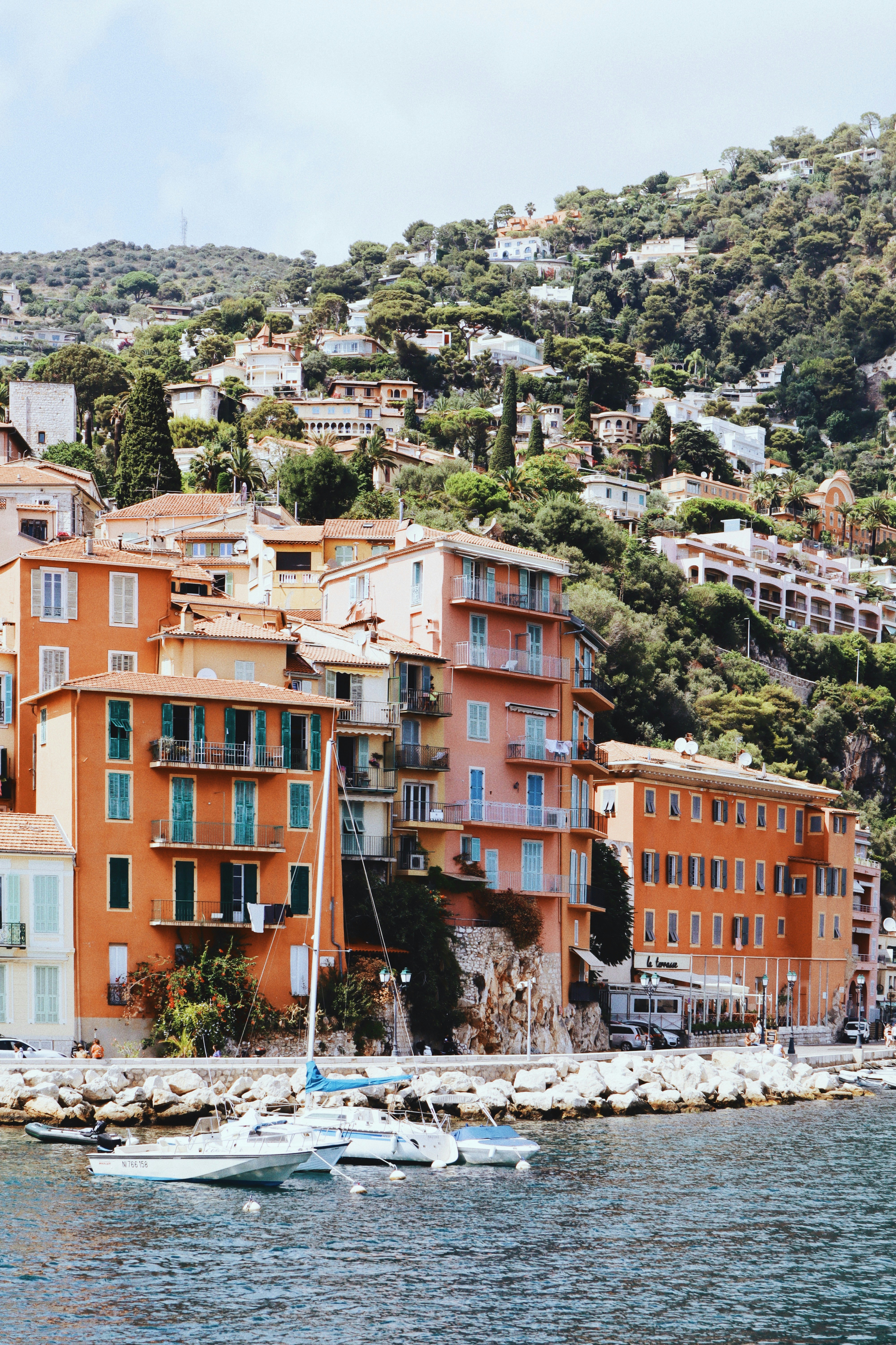 Vibrant waterfront buildings nestled against a lush hillside, showcasing a blend of architecture and natural beauty. A sailboat is anchored in the foreground.