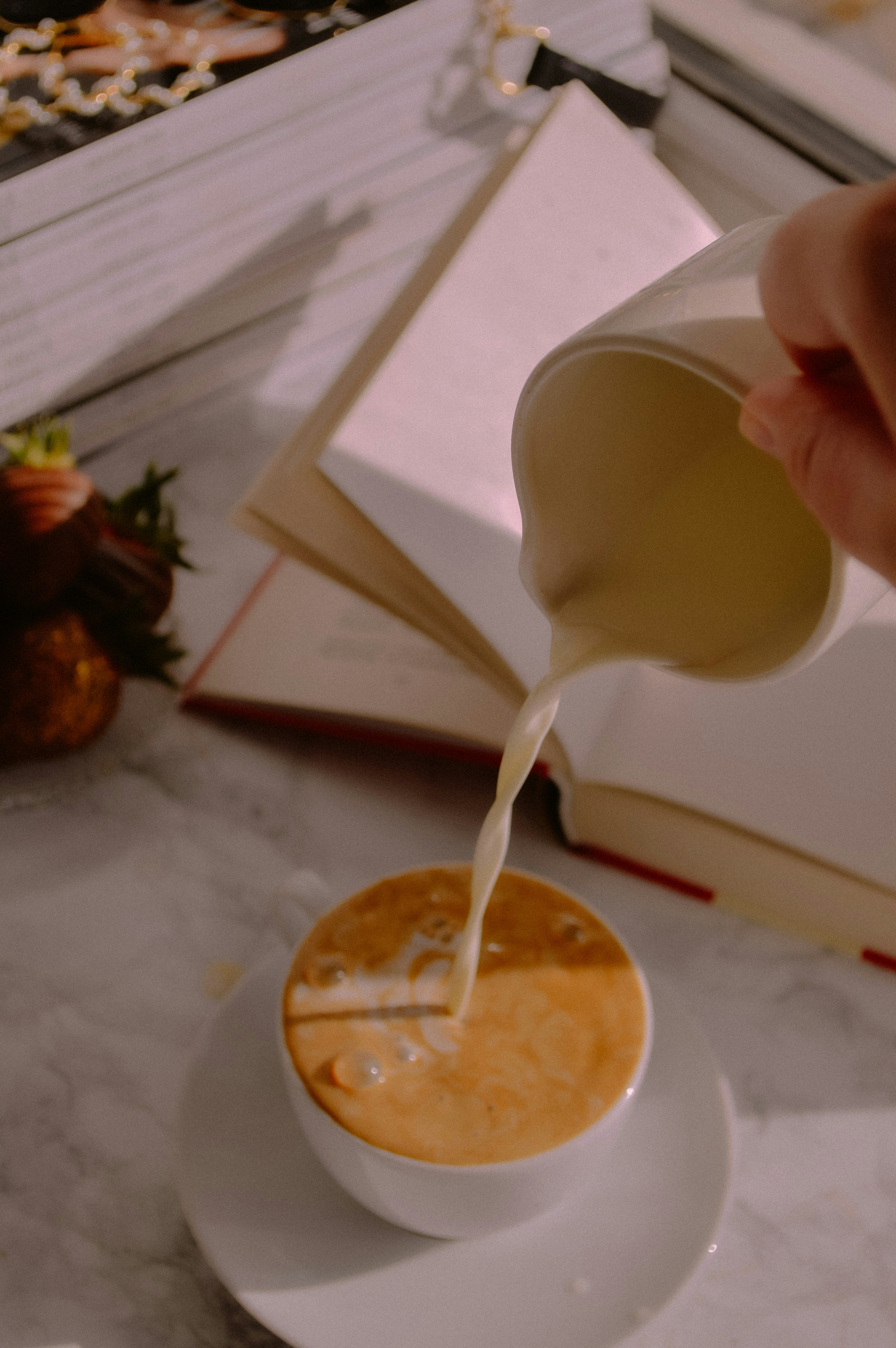Cream being poured into a cup of coffee beside an open book, capturing a cozy moment of relaxation.