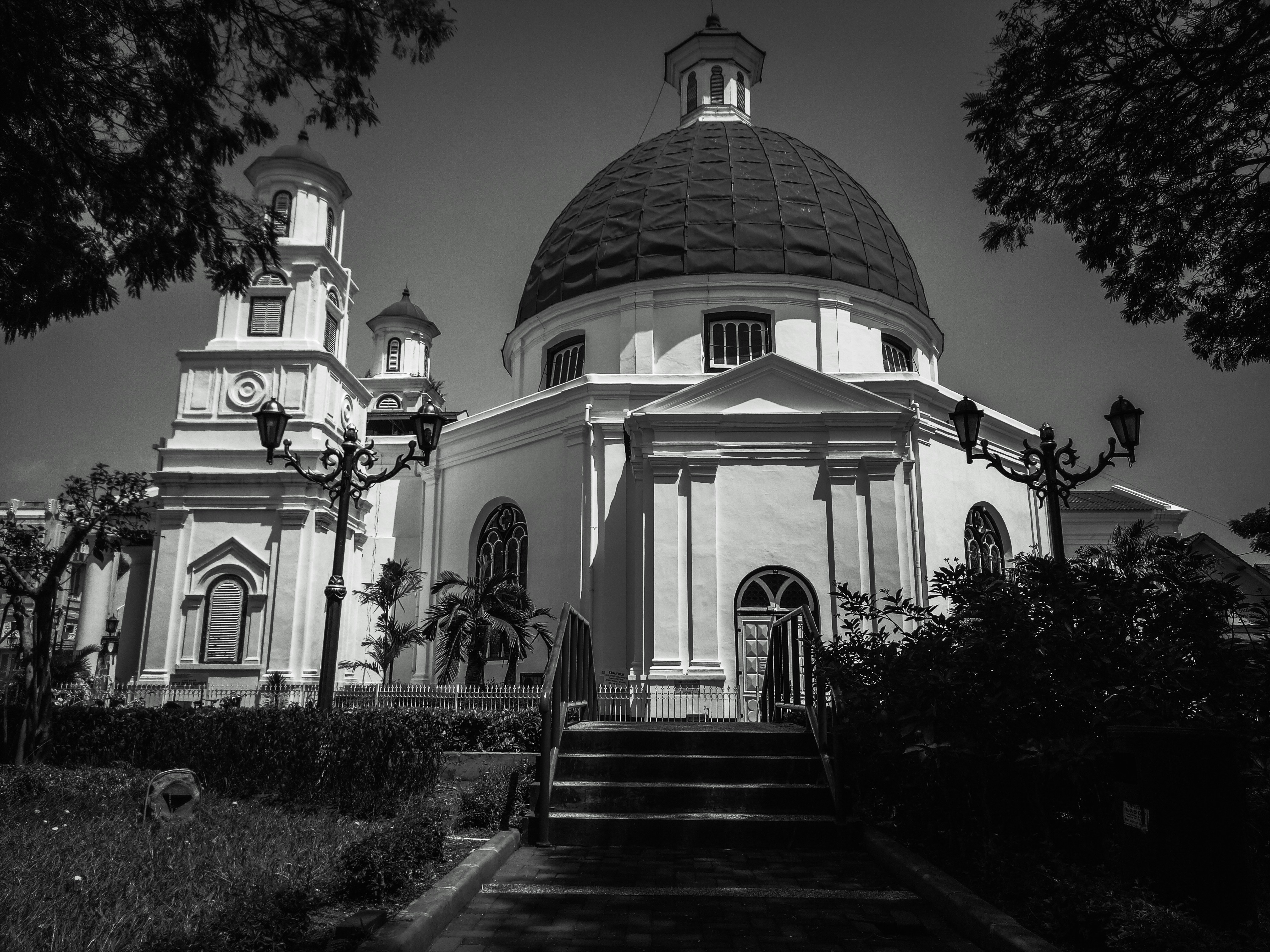 Monochrome façade of a domed church with stairway leading to the door, framed by trees and vintage street lamps.