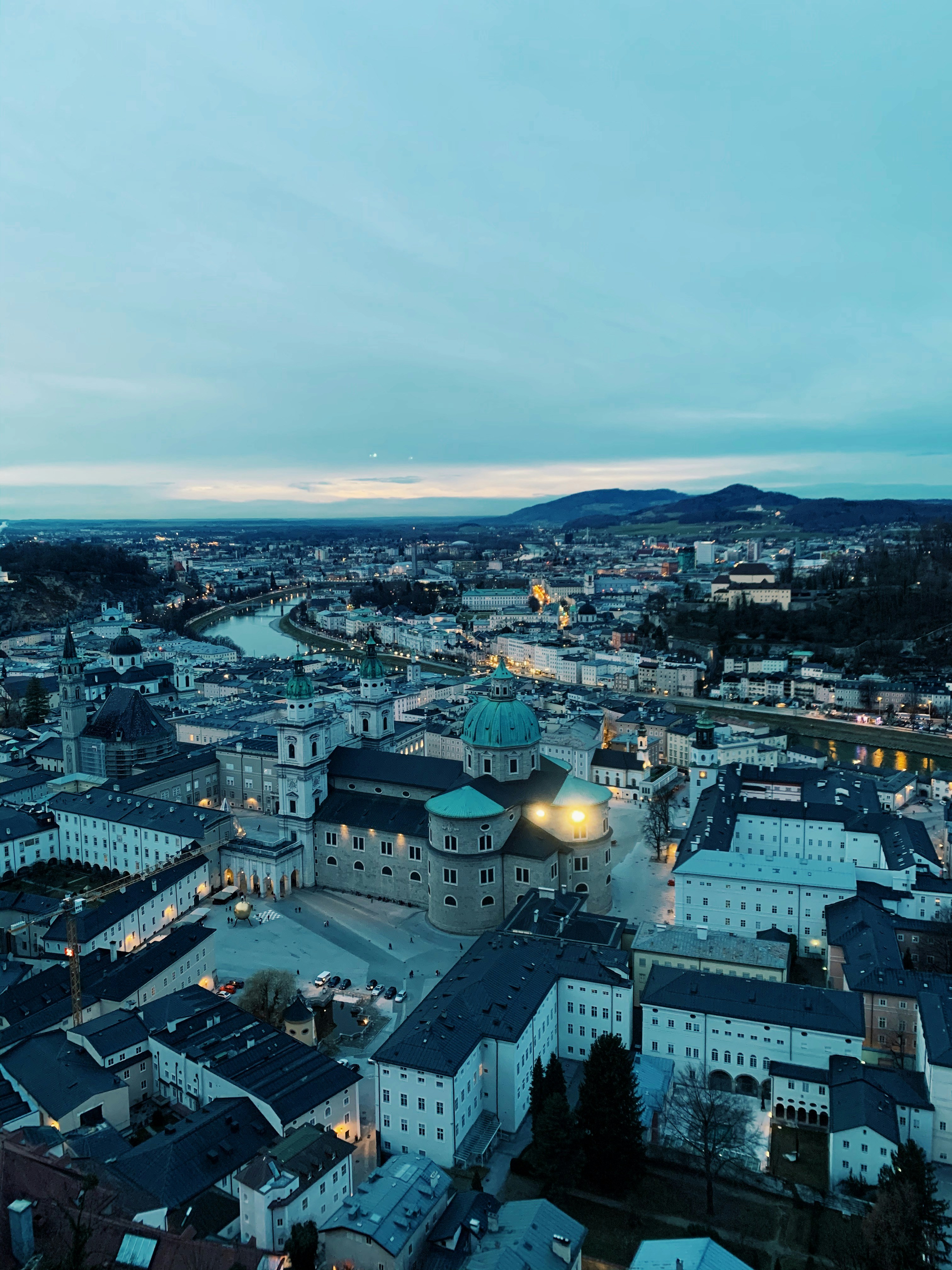 Aerial view of Salzburg showcasing its historic architecture and the winding river at twilight. The scene captures the serene beauty of the city as lights begin to twinkle.